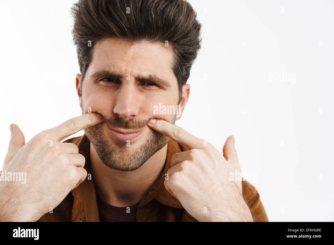 Unhappy young unshaven man pointing fingers at his cheeks isolated over ...