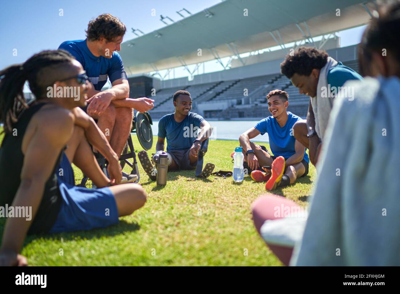 Young male athlete friends resting and talking on sunny stadium lawn ...