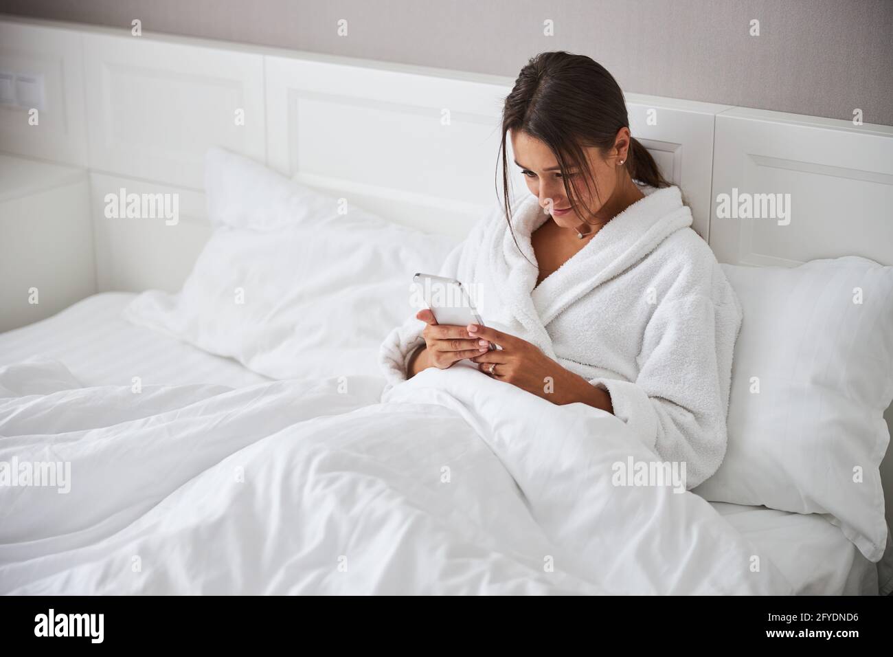 Beautiful Caucasian female in white bathrobe lying on bed while using ...