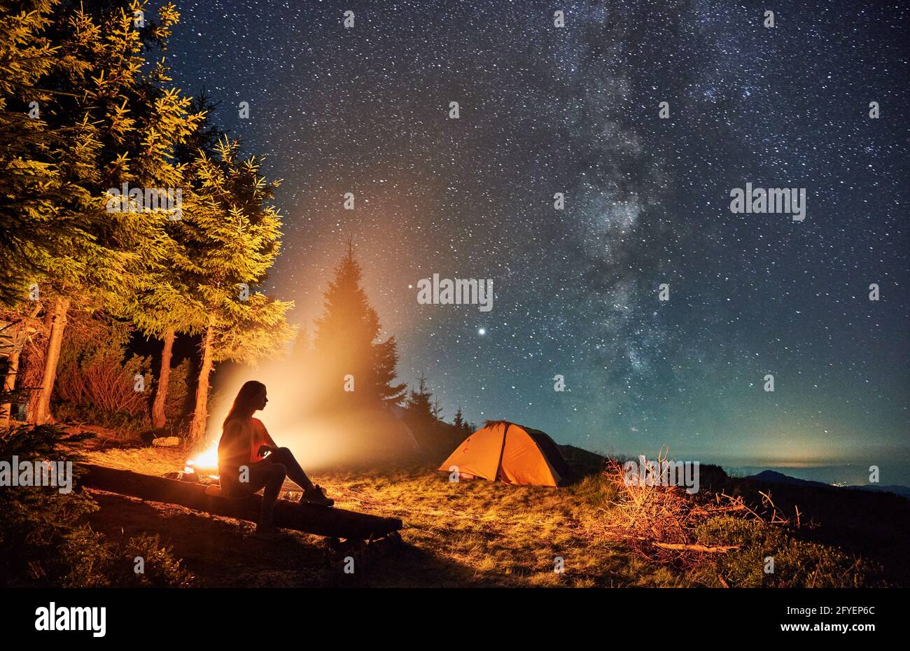Young woman hiker sitting on bench near bonfire under magical sky with ...
