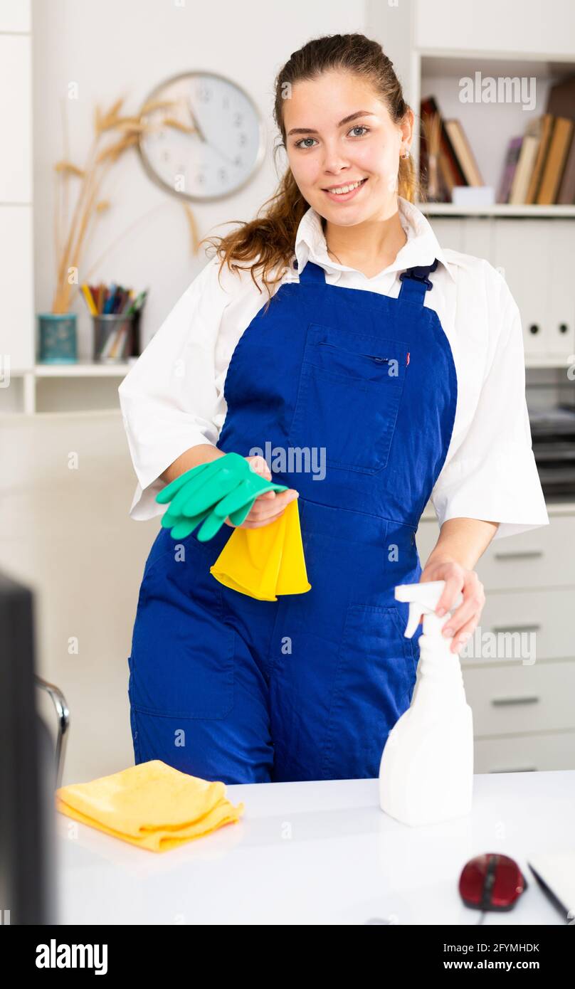 Young friendly cleaning woman in work uniform removes dust with a spray ...