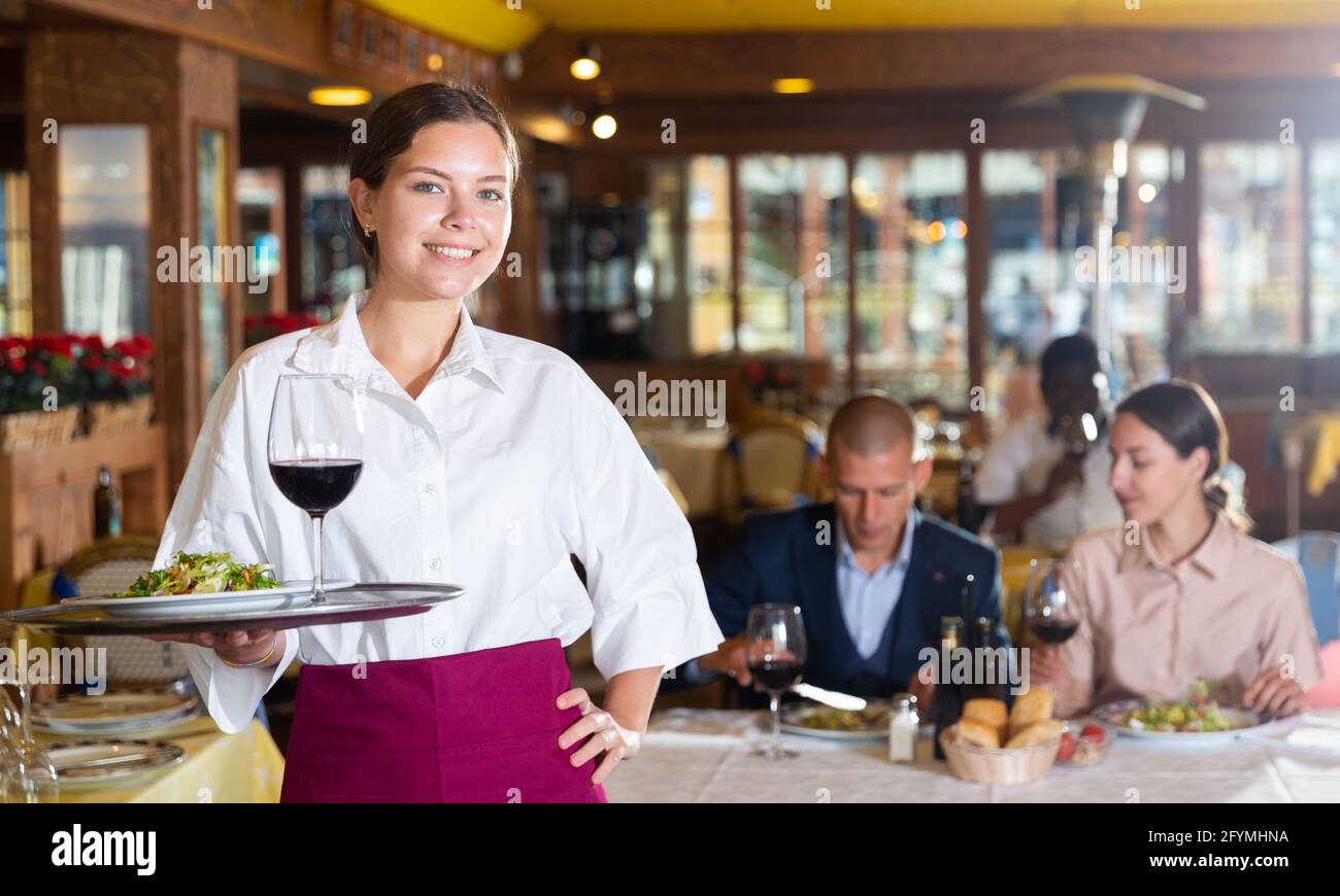 Polite waiter holding tray at restaurant with customers behind him ...