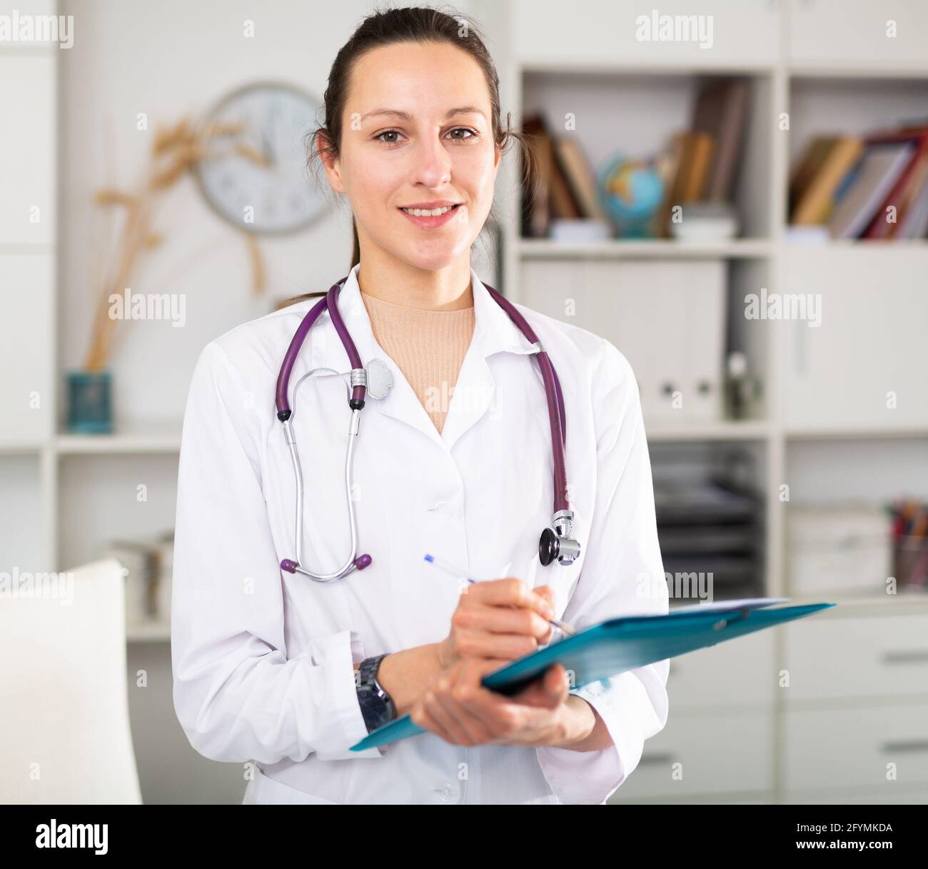 Positive woman doctor in formal wear standing with folder in office ...