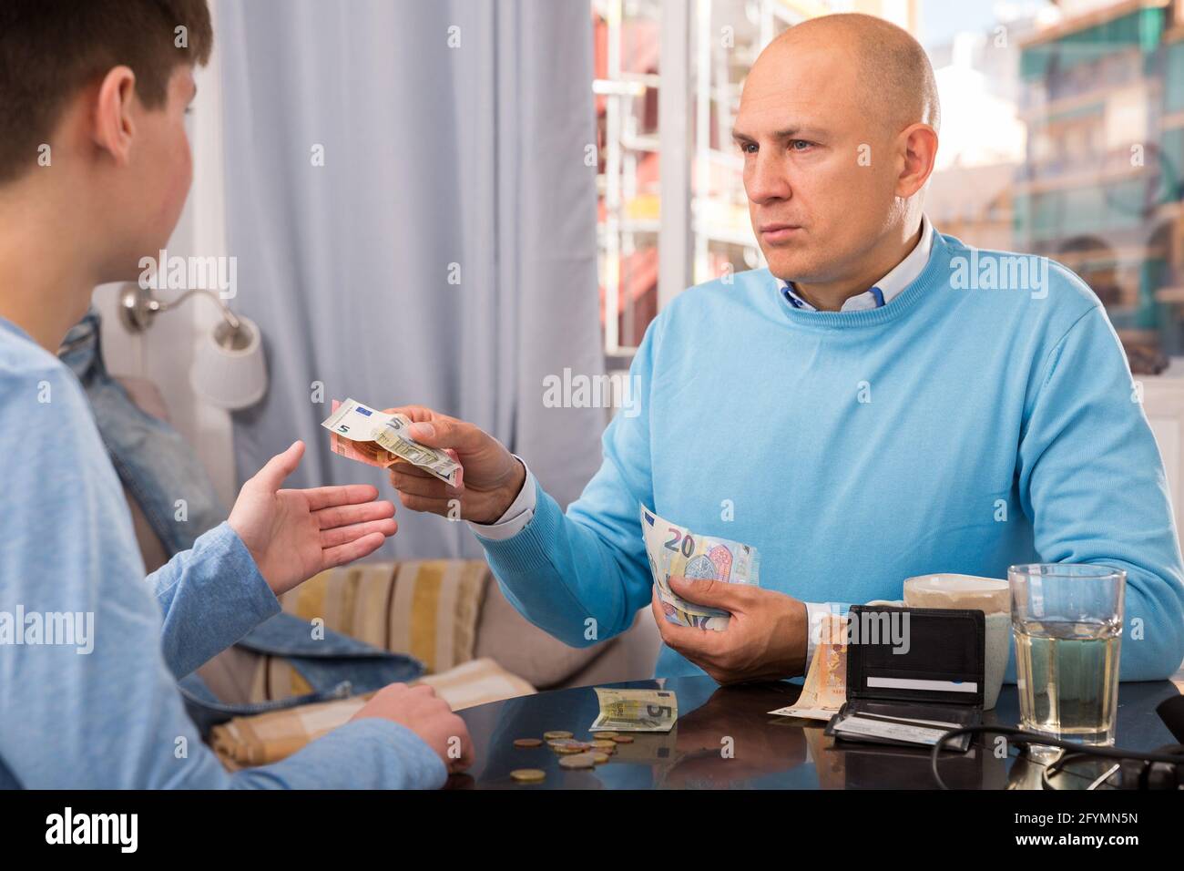 Portrait of positive man giving money to teen boy in home interior ...