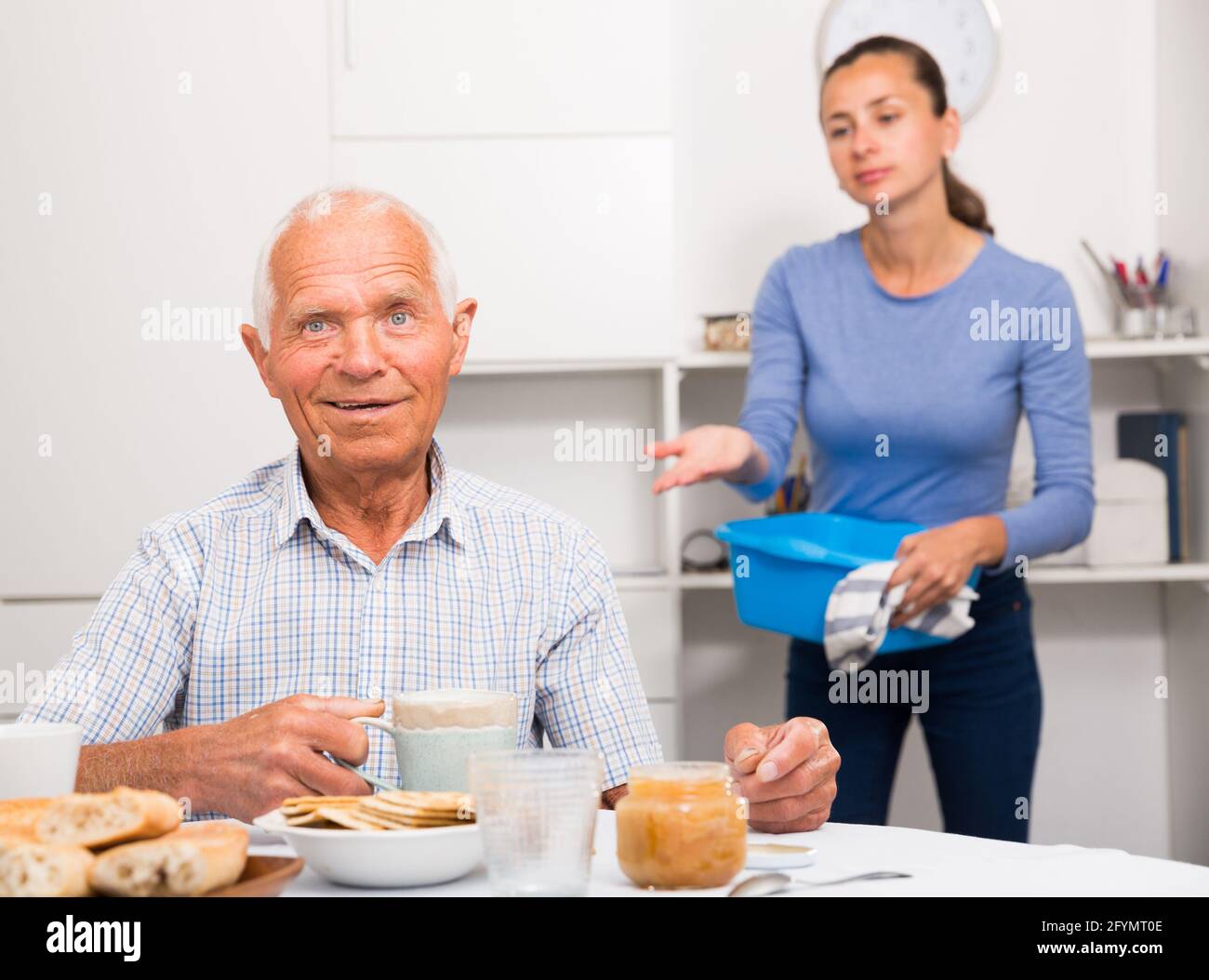 Mature father drinking tea in the kitchen. Adult daughter does cleaning ...