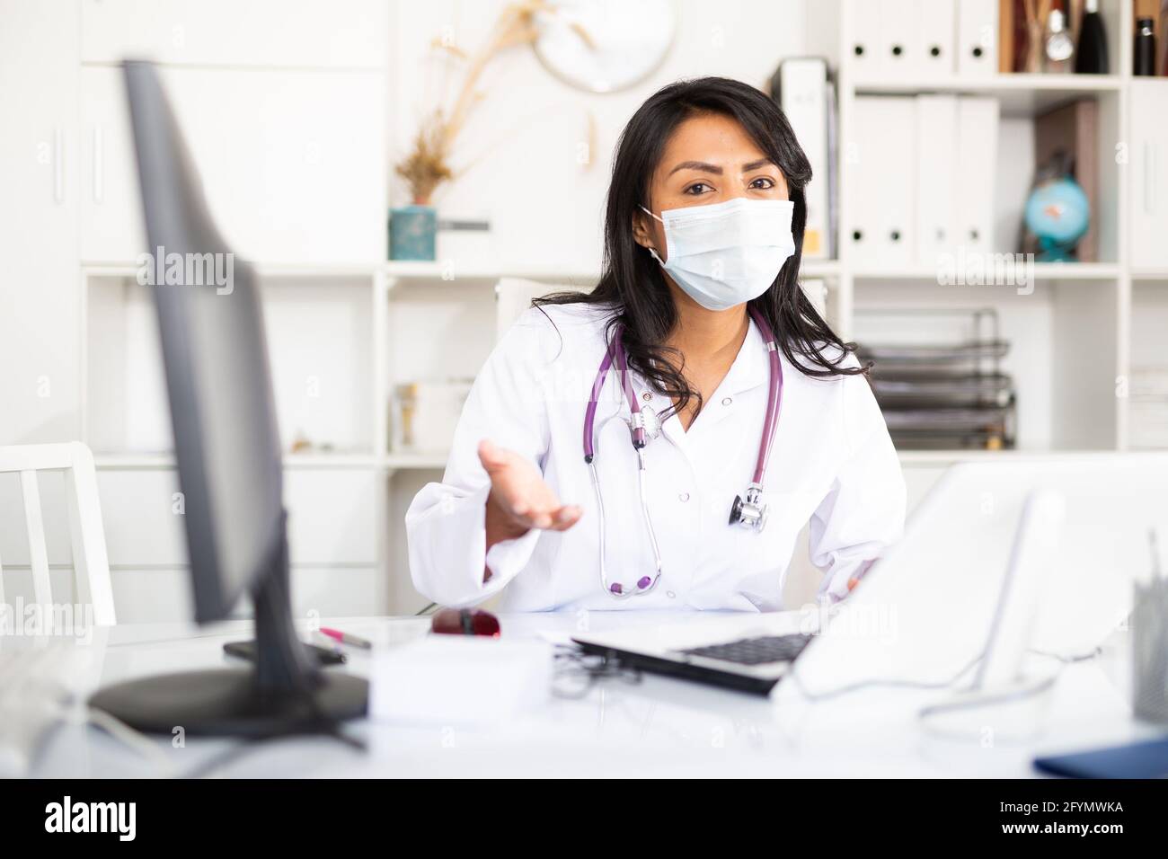 Female doctor wearing surgical mask sitting at table in medical office ...