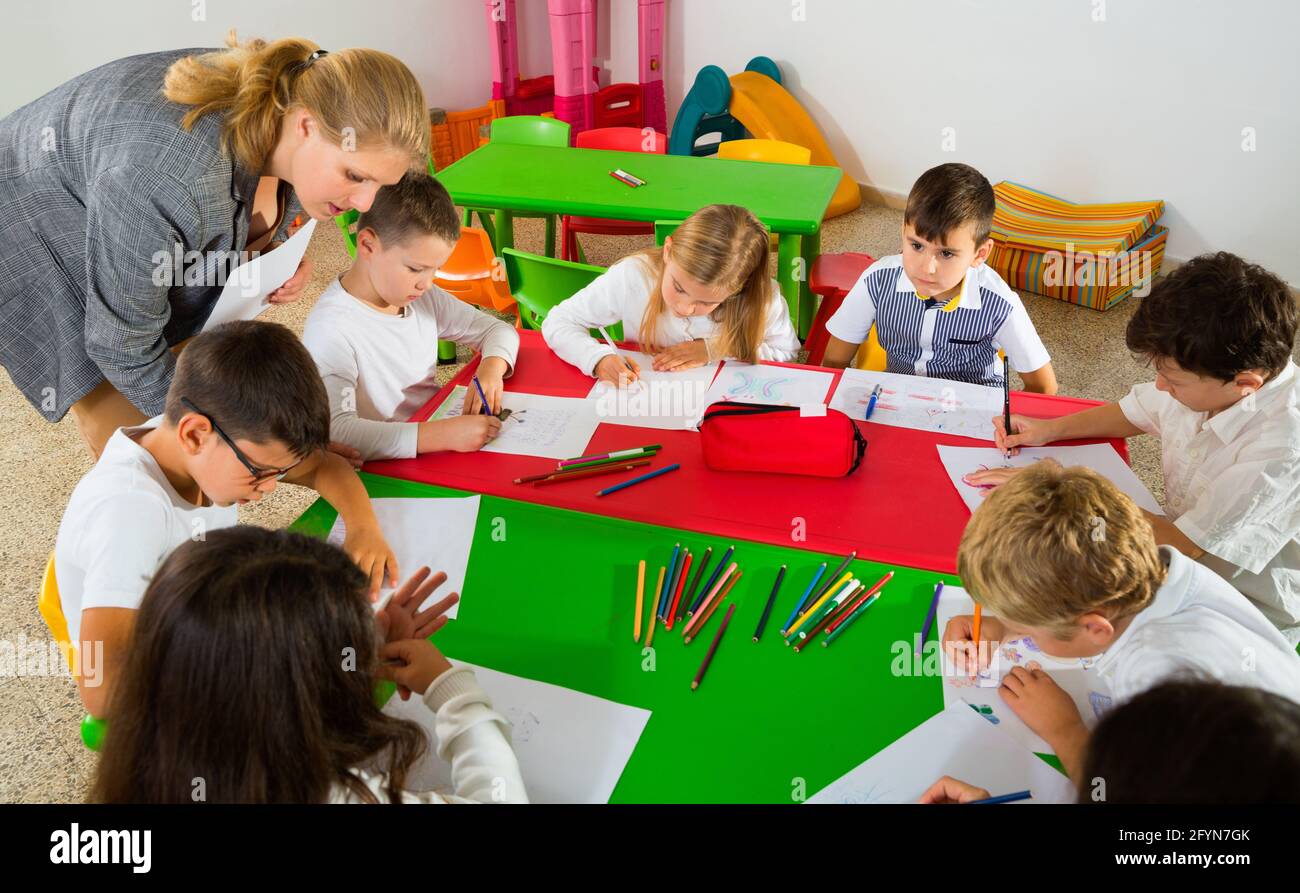 Cheerful smiling female teacher helping schoolkids drawing with color ...