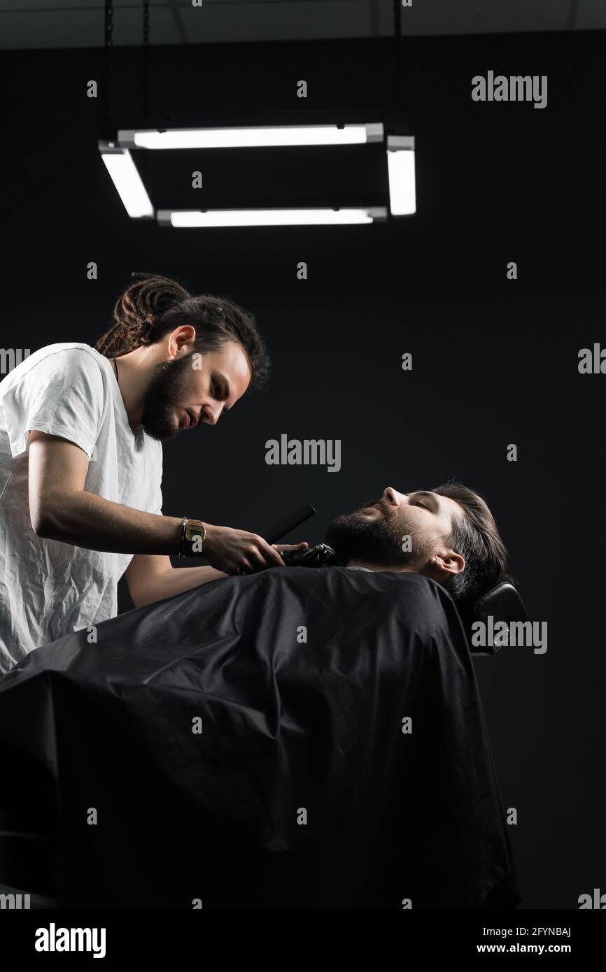 Beard shaving in barbershop. Barber with dreadlocks trim handsome ...