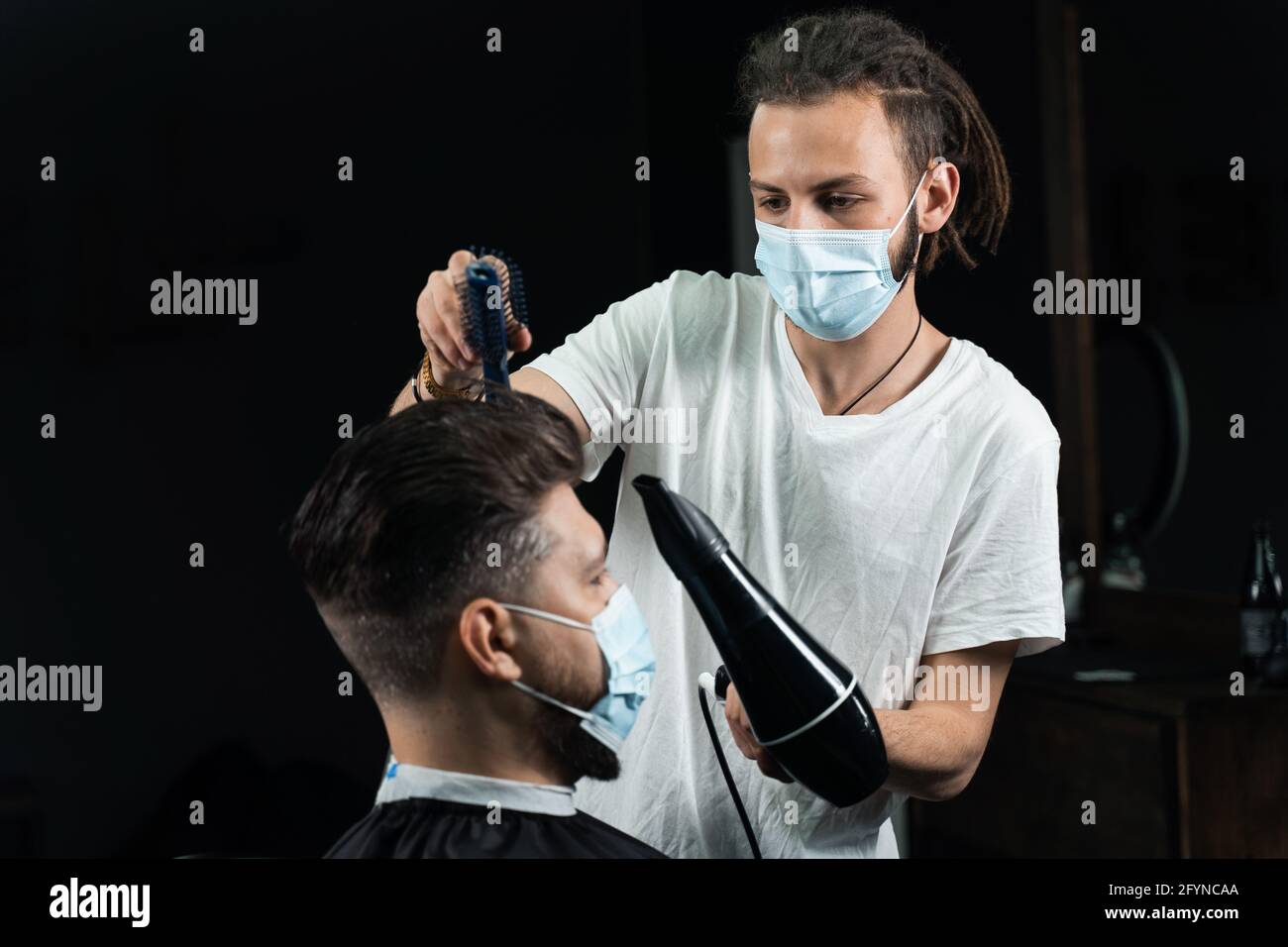 Barber in medical mask dries the hair of a handsome bearded man after a ...