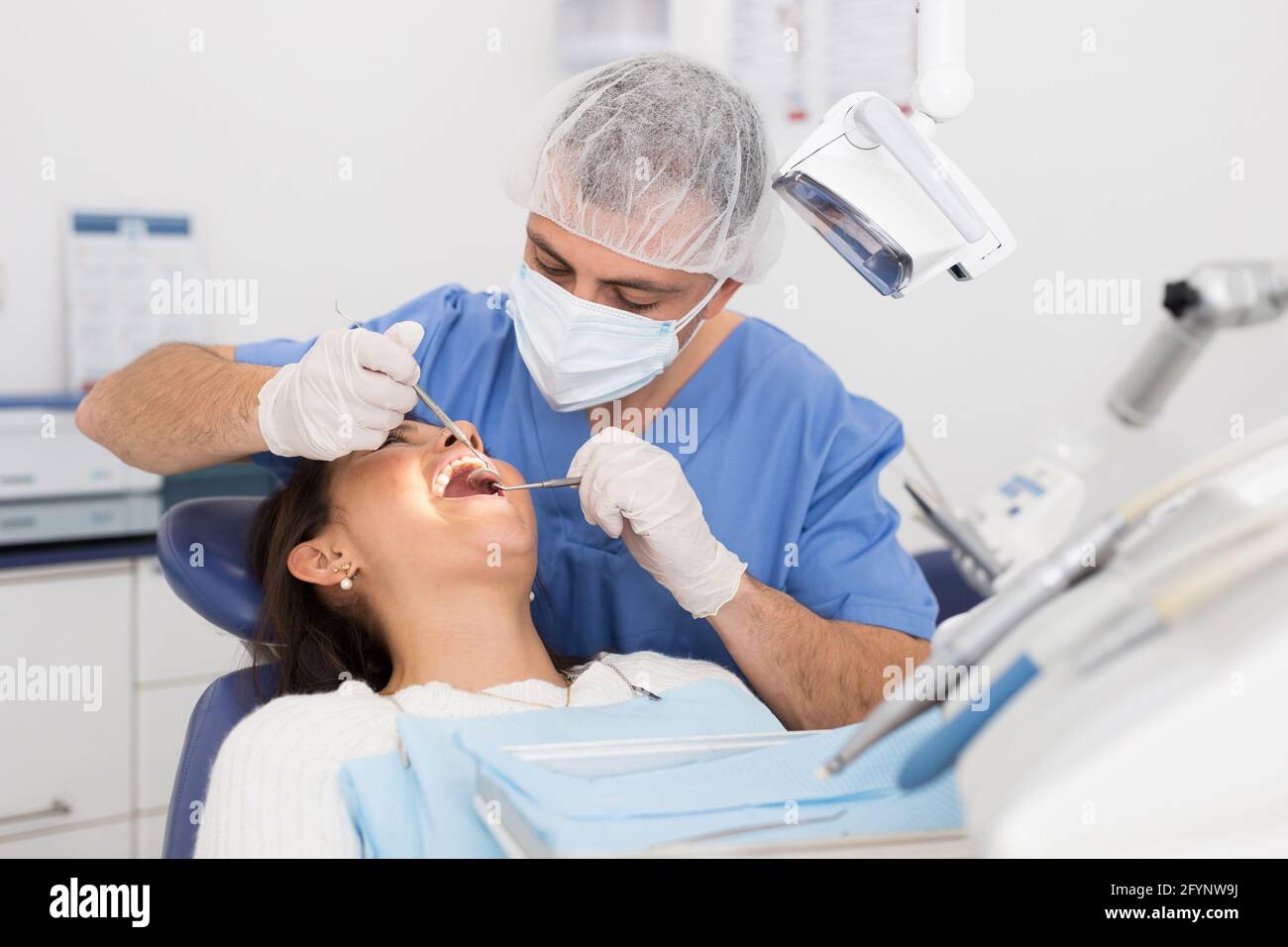 The dentist in face mask inspects female patient teeth with mirror and ...