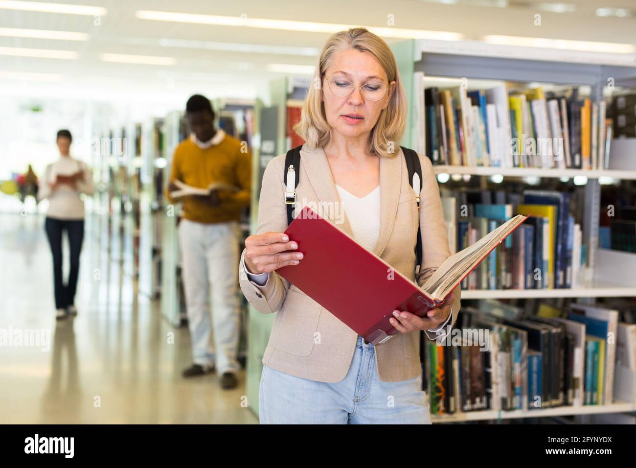 Mature woman reading book while standing near bookshelves in public ...