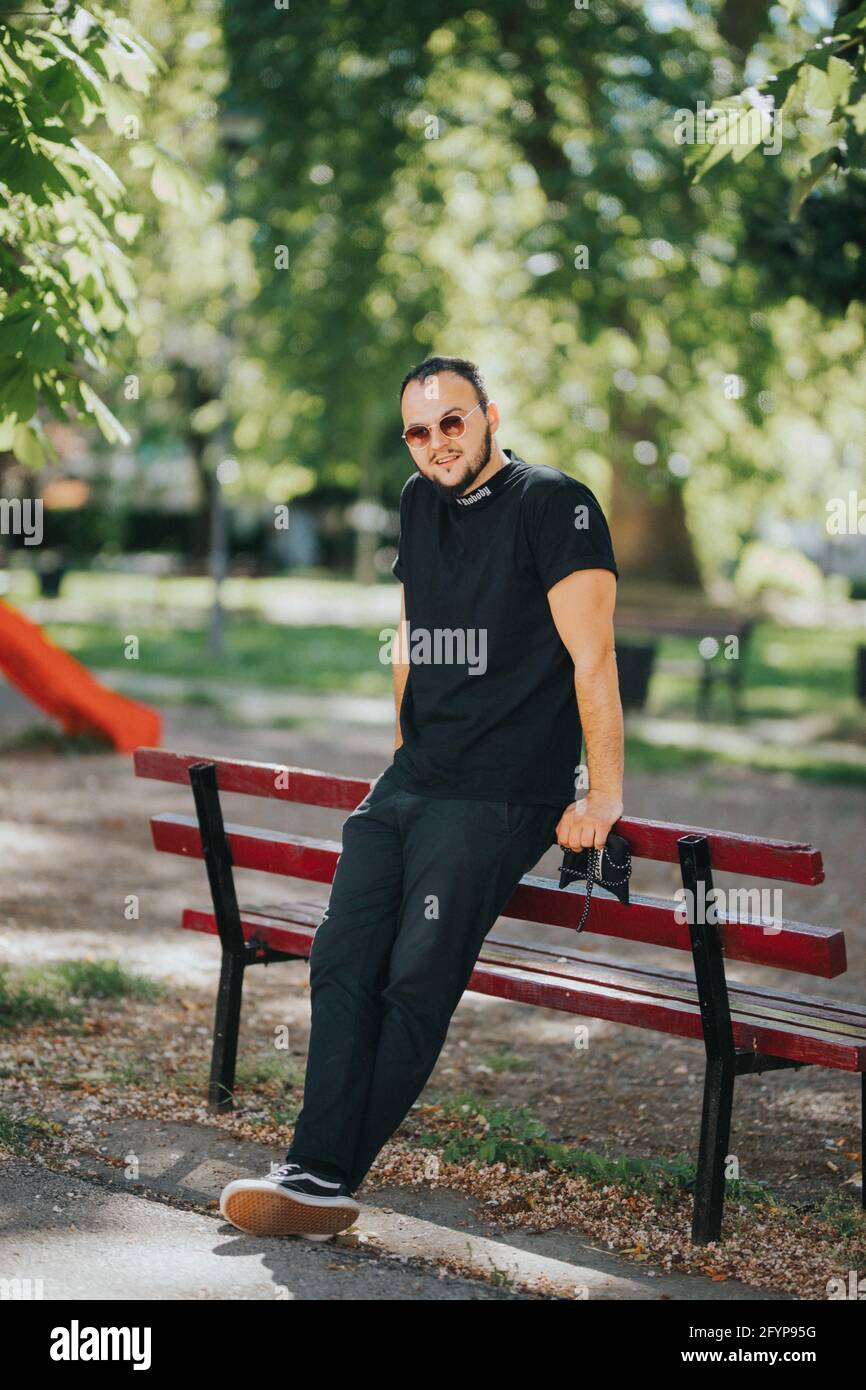 Handsome guy leaning on a wooden bench is posing in the summer park ...