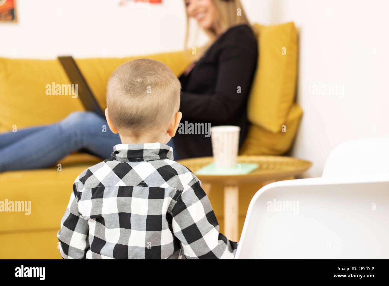 A cute little boy looking at her mother while she is working via laptop ...