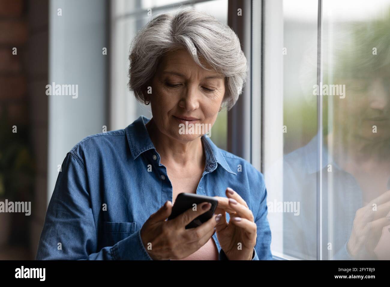 Serious mature woman standing near window with smartphone Stock Photo ...