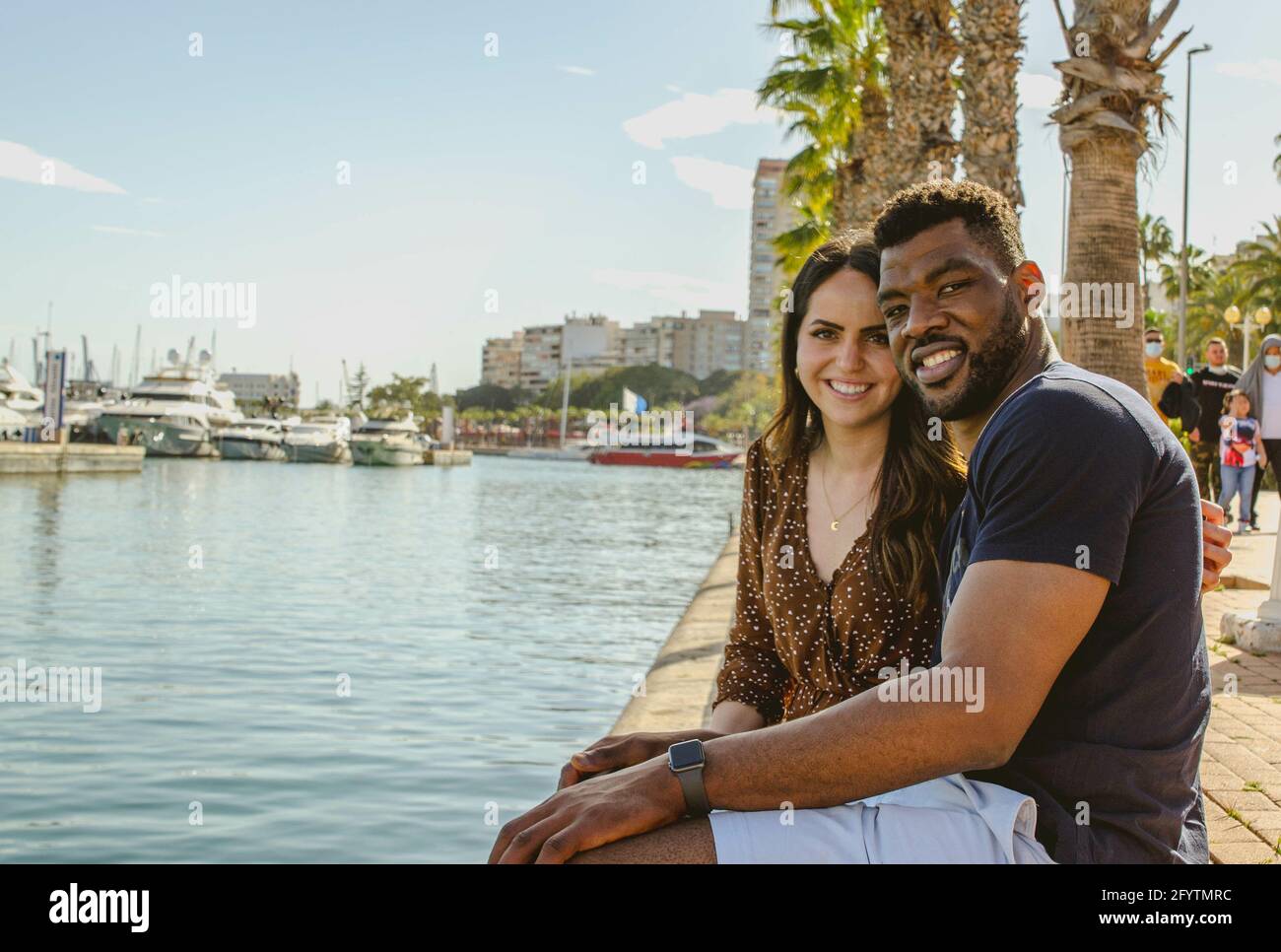 Picture of Young Adult Interracial Couple Smiling Looking at Camera Sitting on Harbor Port With ...