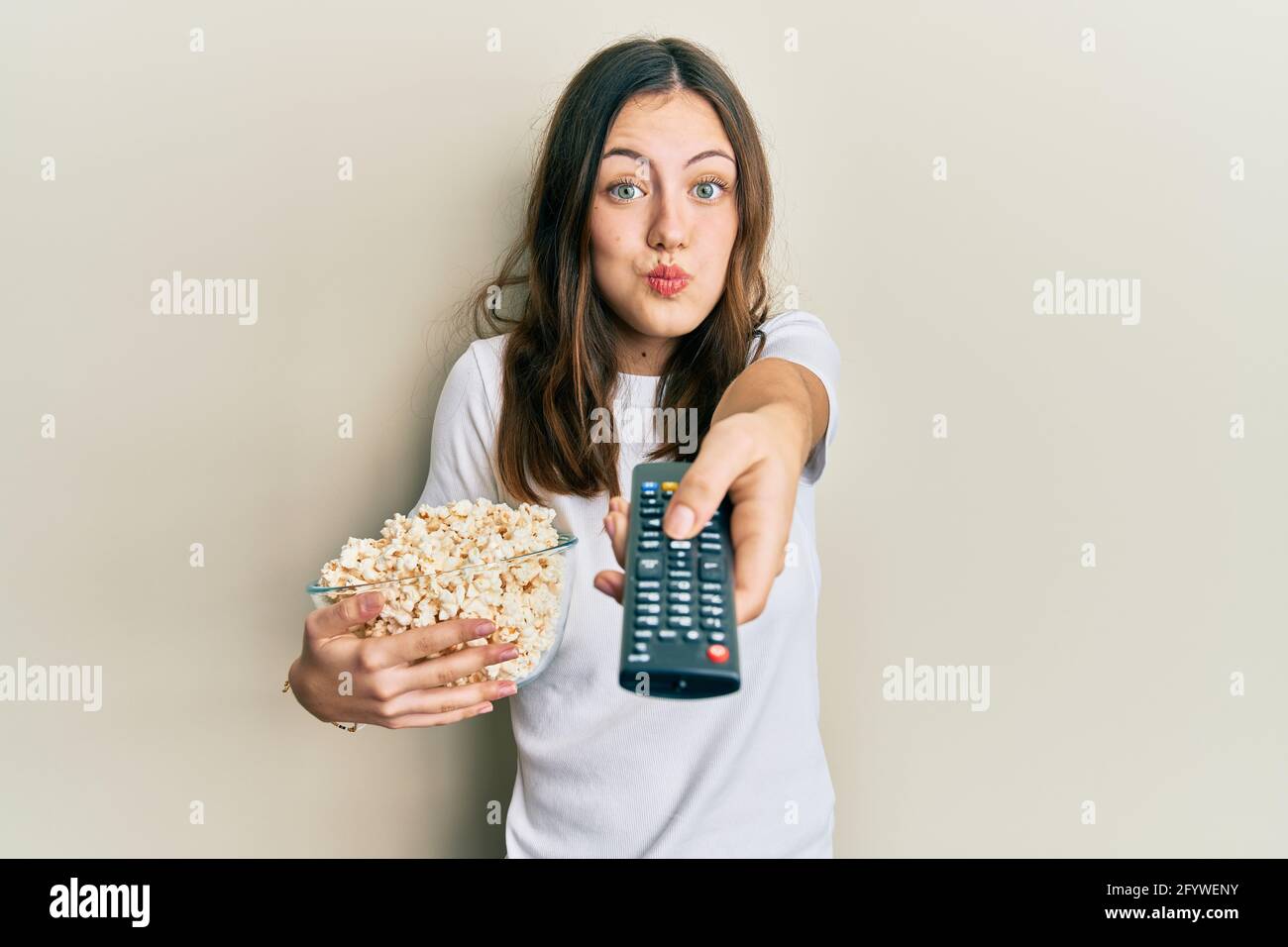 Young brunette woman eating popcorn using tv control puffing cheeks ...