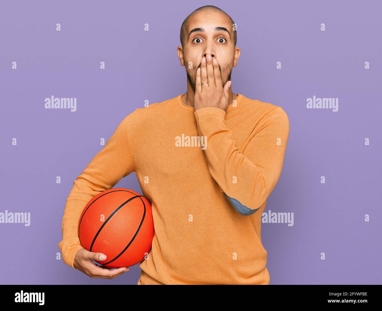 Hispanic adult man holding basketball ball covering mouth with hand ...