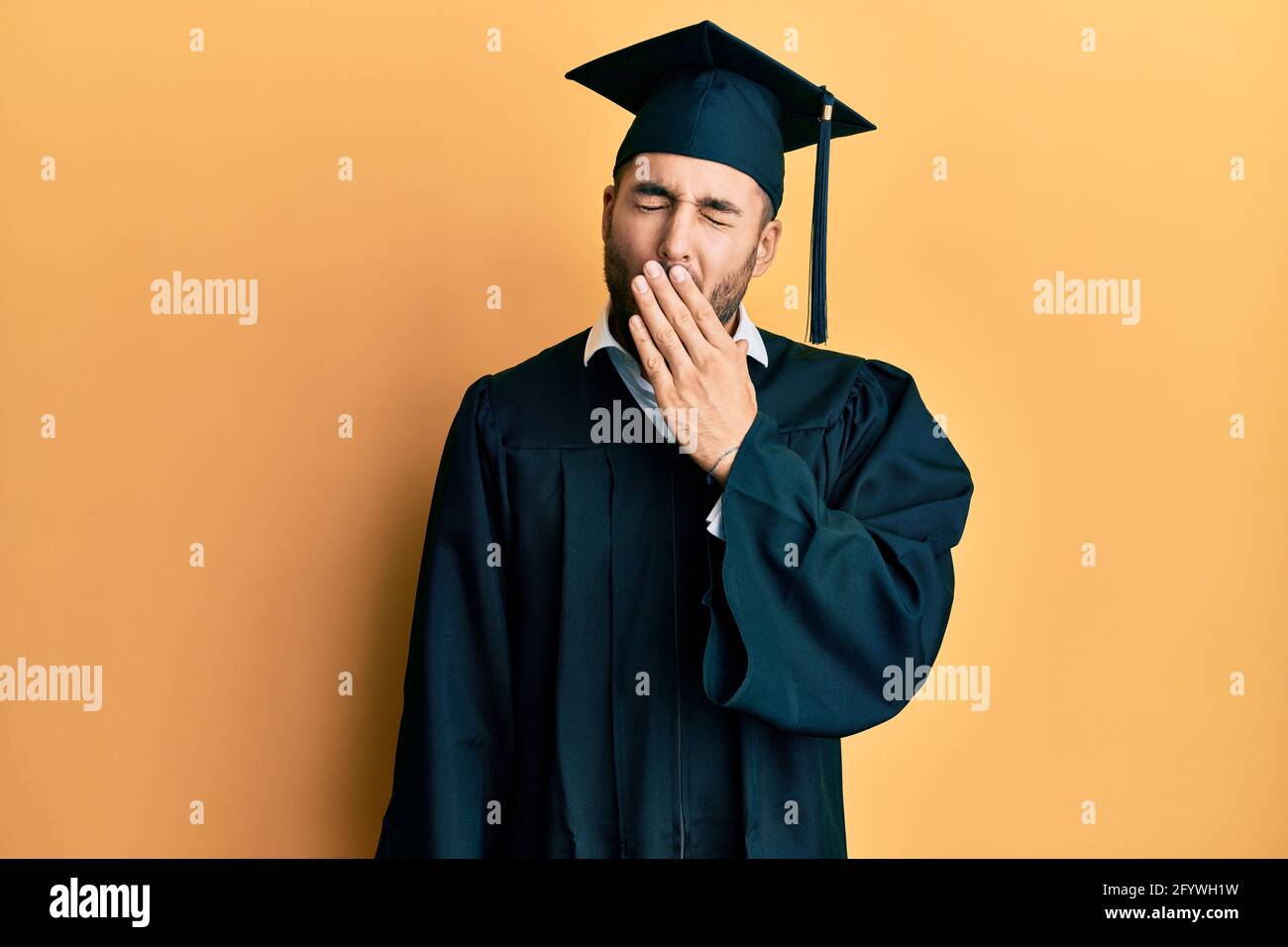 Young hispanic man wearing graduation cap and ceremony robe bored ...