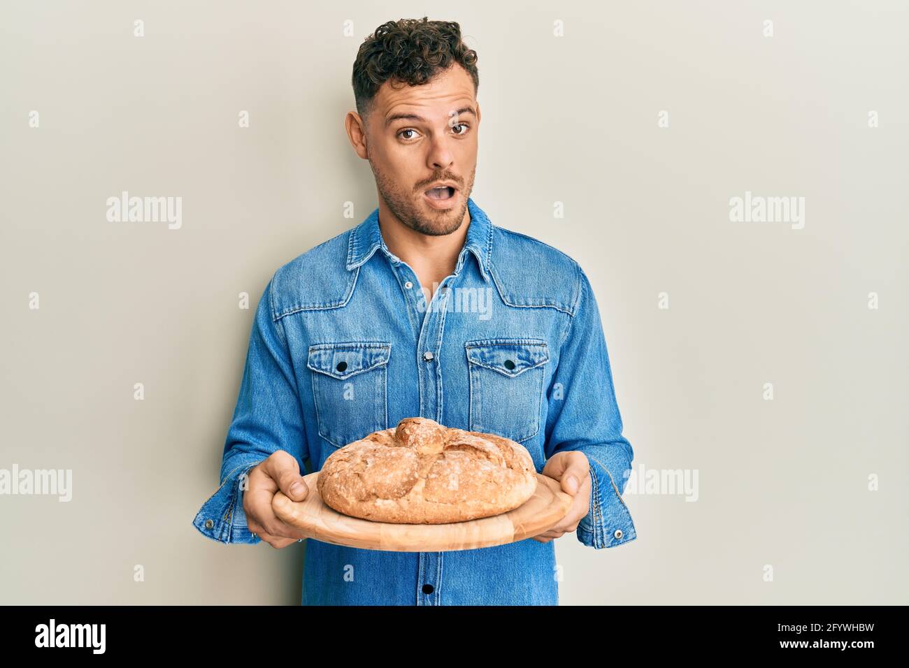 Young hispanic man holding bread in shock face, looking skeptical and ...