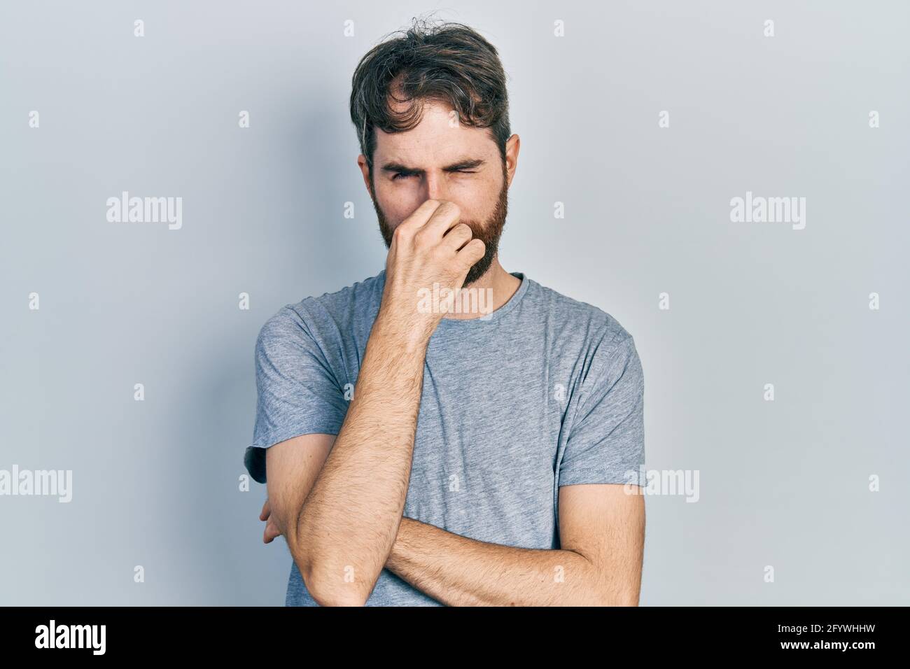 Caucasian man with beard wearing casual grey t shirt smelling something ...