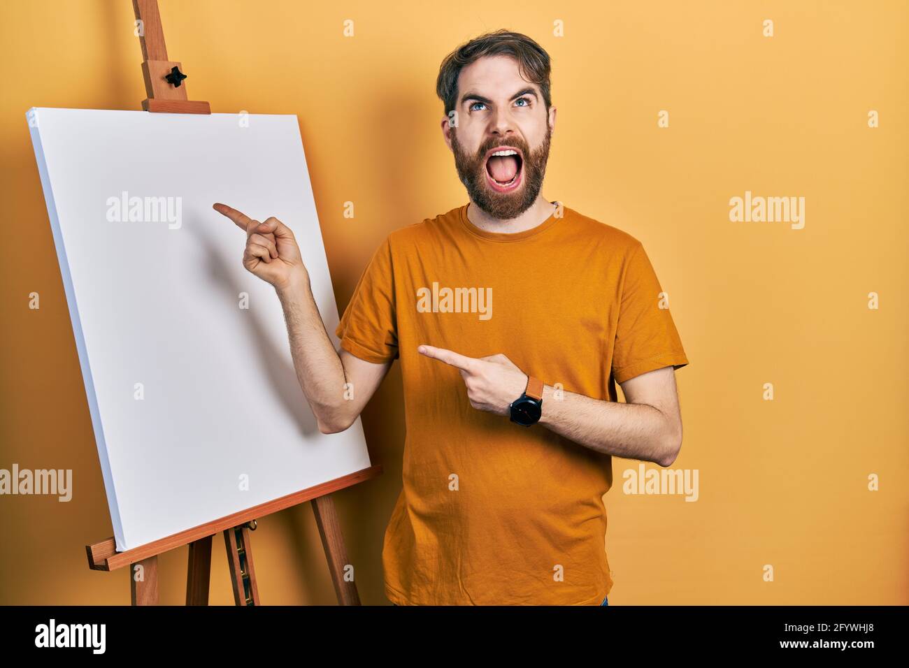 Caucasian man with beard pointing to painter easel stand angry and mad ...