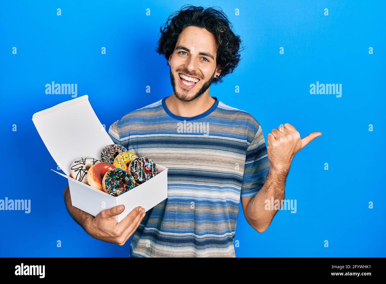 Handsome hispanic man holding tasty colorful doughnuts pointing thumb ...