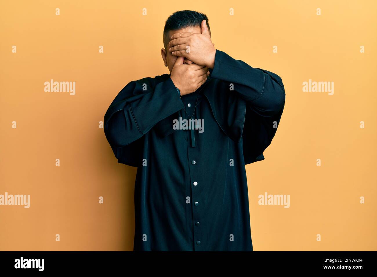 Young latin priest man standing over yellow background covering eyes ...