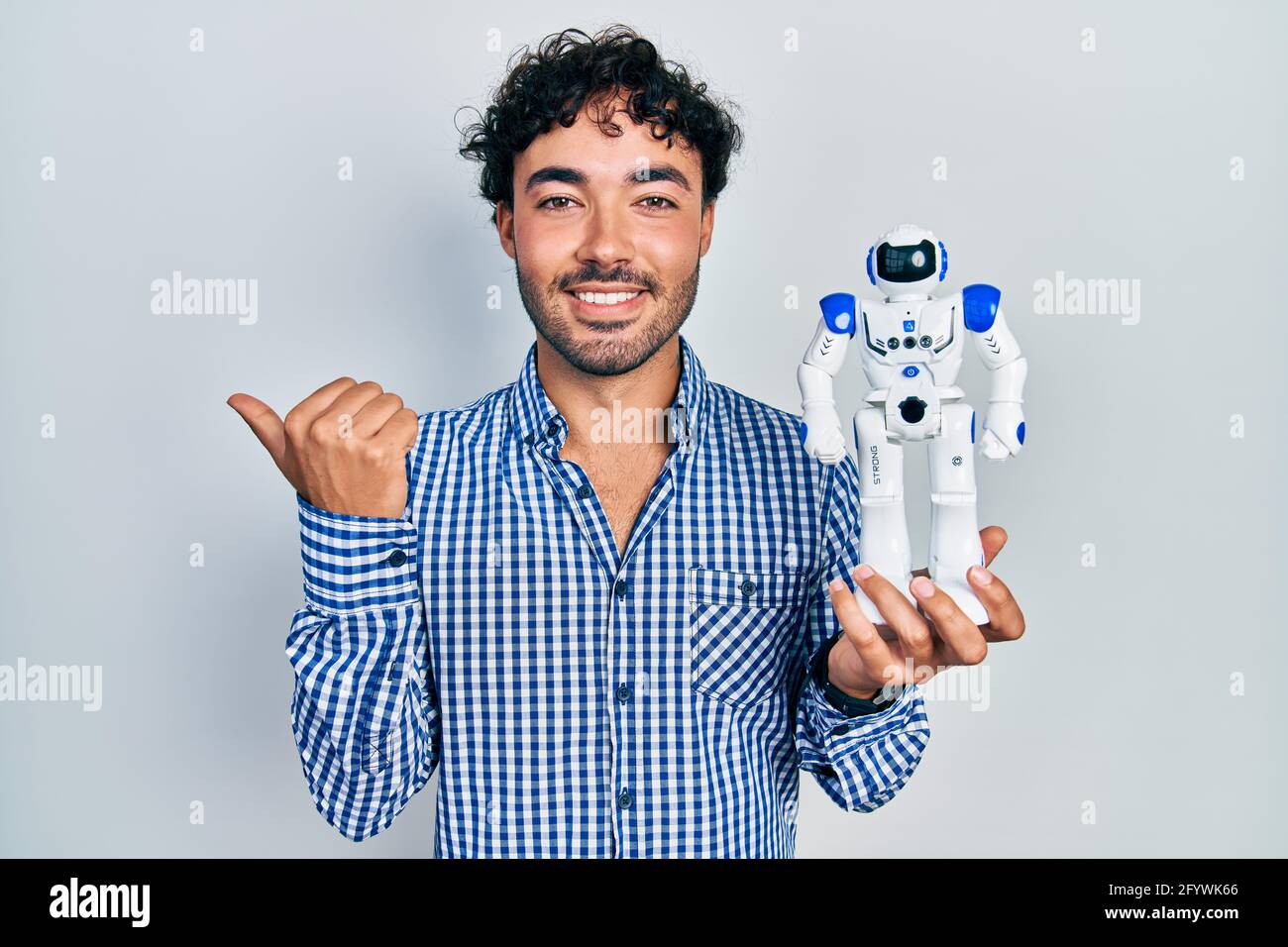 Young hispanic man holding robot toy pointing thumb up to the side ...