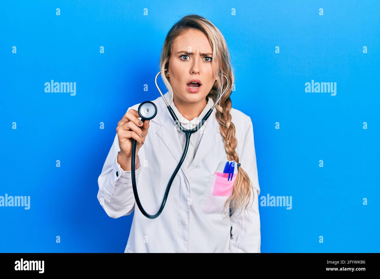 Beautiful young blonde doctor woman holding stethoscope in shock face ...
