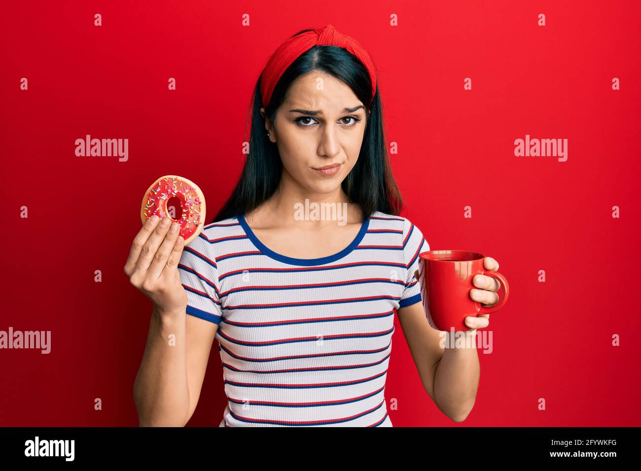 Young hispanic woman eating doughnut and drinking coffee skeptic and ...