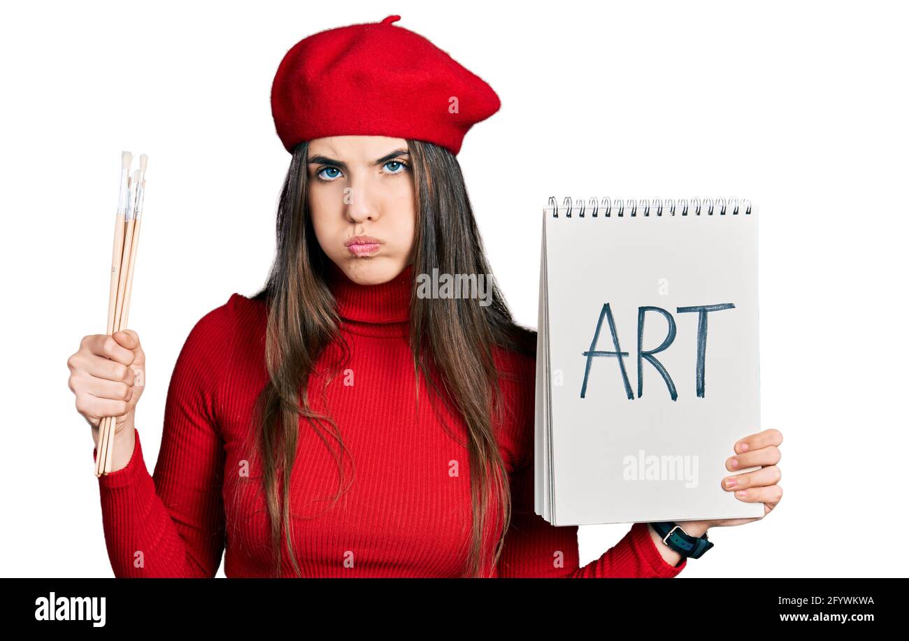 Young brunette teenager holding art notebook and paintbrushes puffing ...