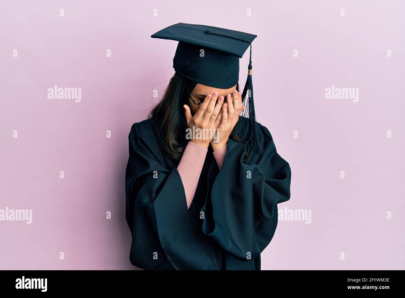 Young hispanic woman wearing graduation cap and ceremony robe with sad ...