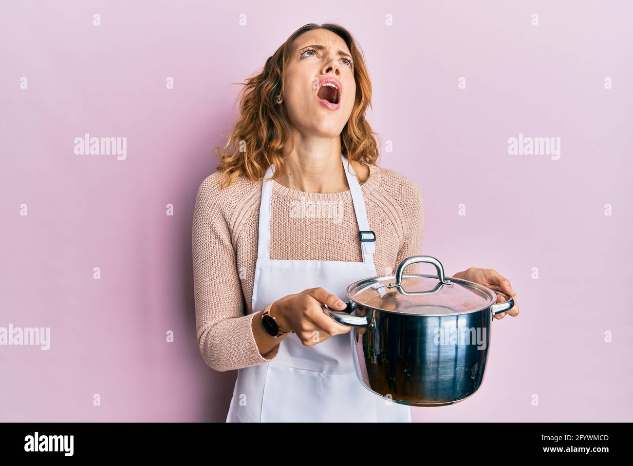 Young caucasian woman wearing apron holding cooking pot angry and mad ...