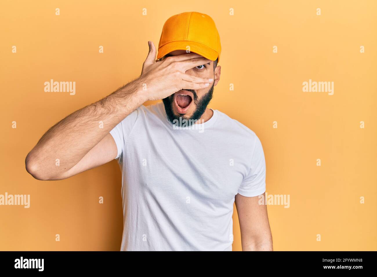 Young man with beard wearing yellow cap peeking in shock covering face ...