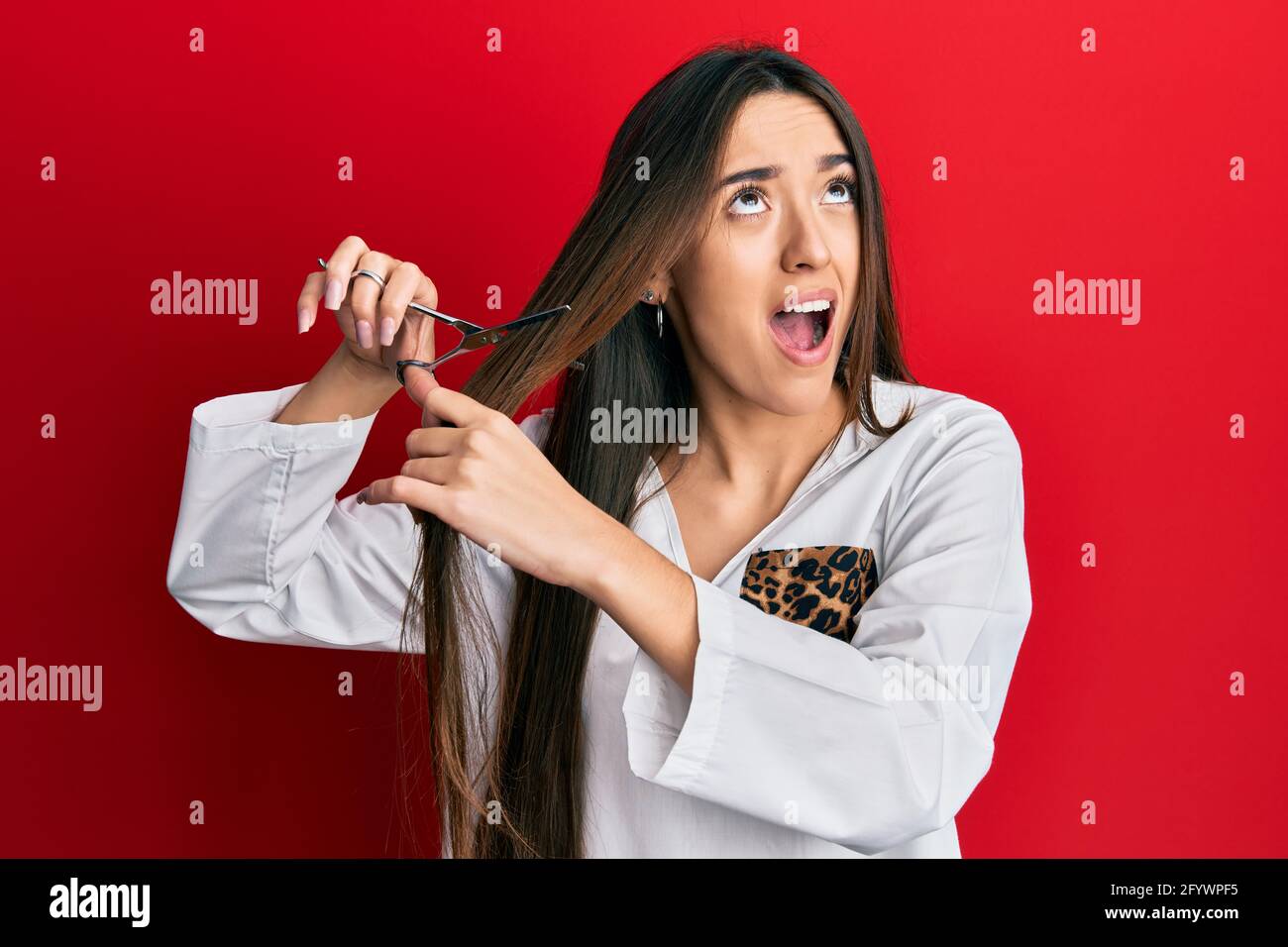 Young hispanic girl cutting hair using scissors angry and mad screaming frustrated and furious ...