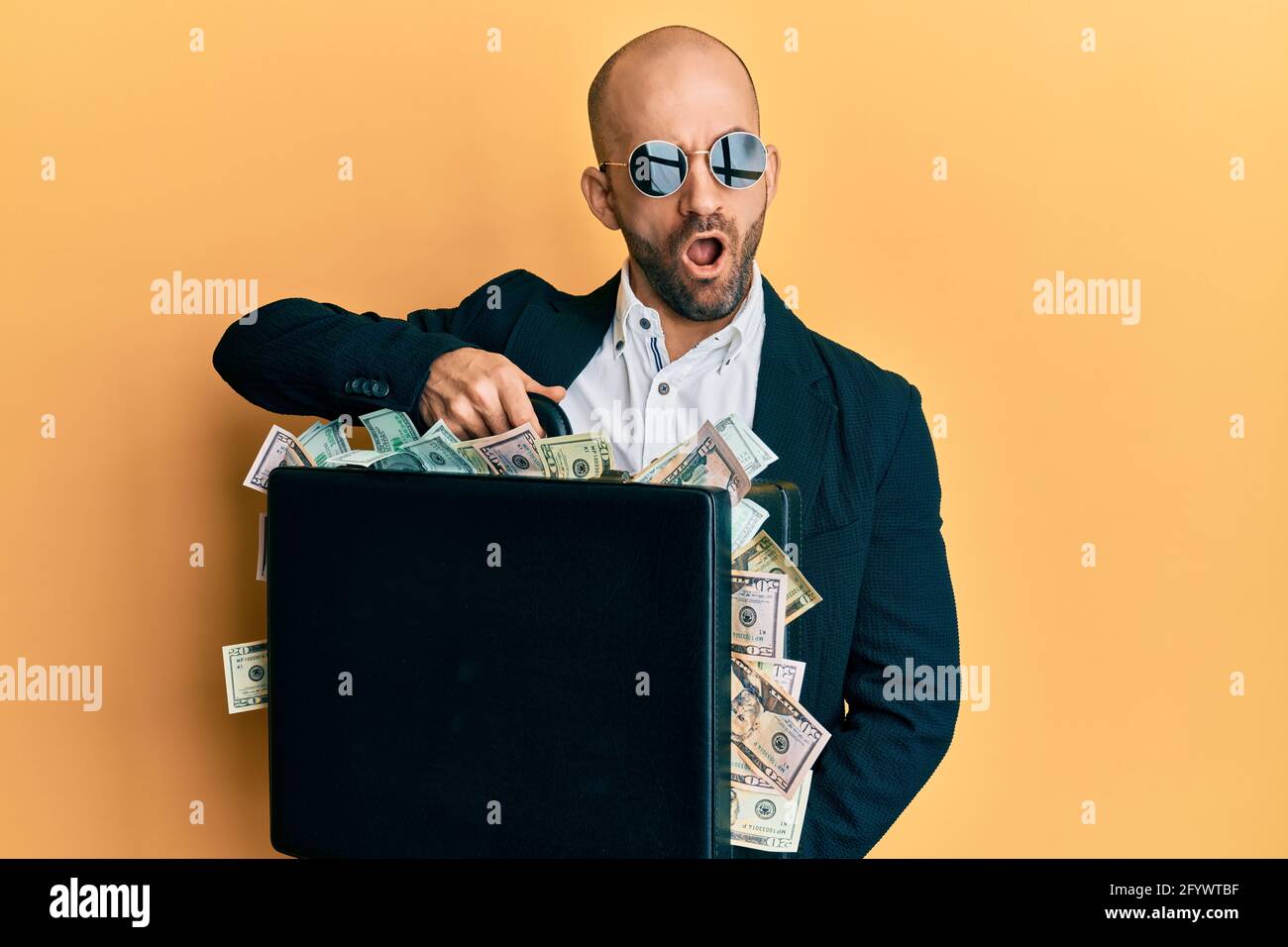 Young hispanic man holding briefcase with dollars in shock face ...