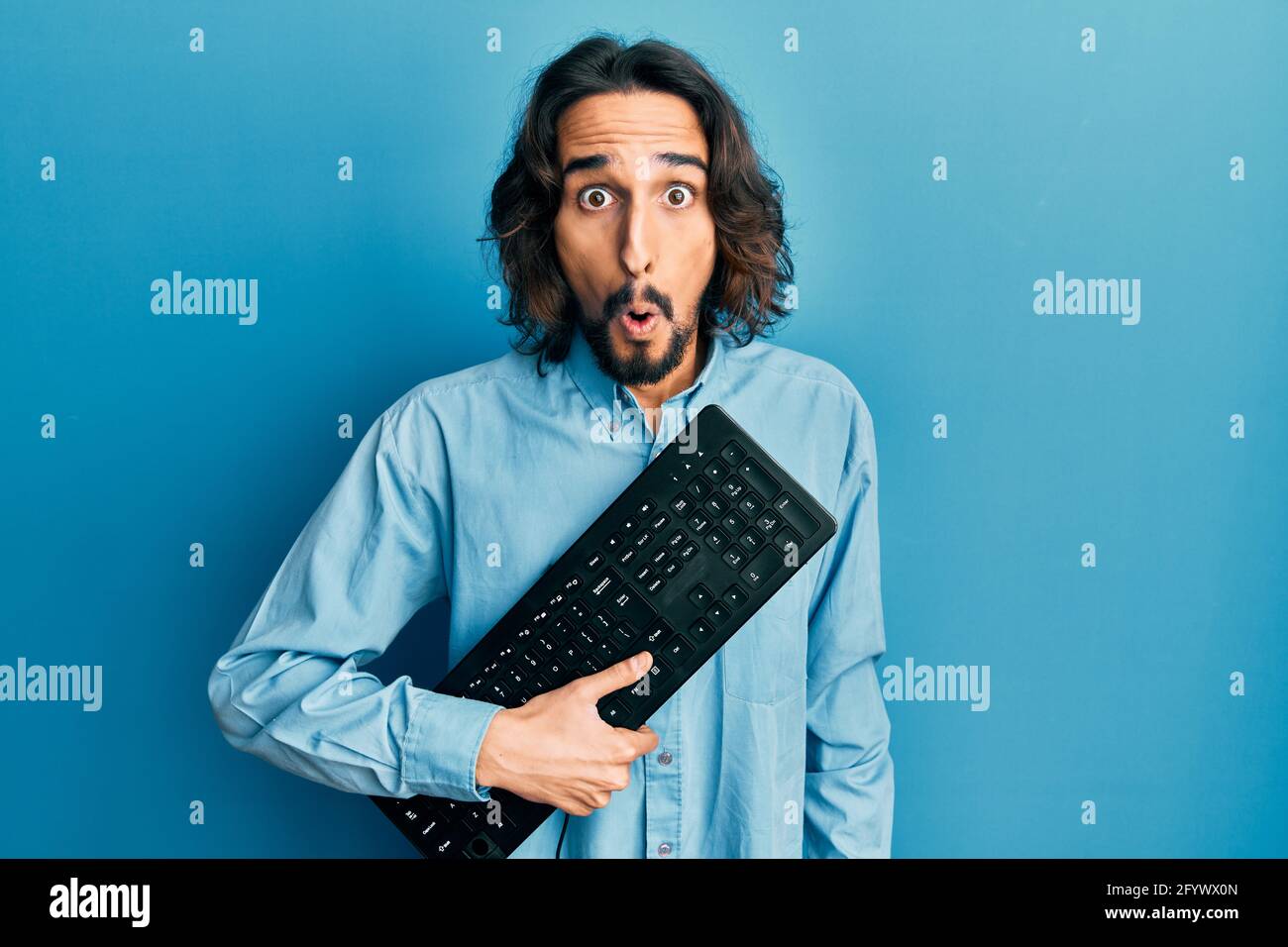 Young hispanic man holding keyboard scared and amazed with open mouth ...