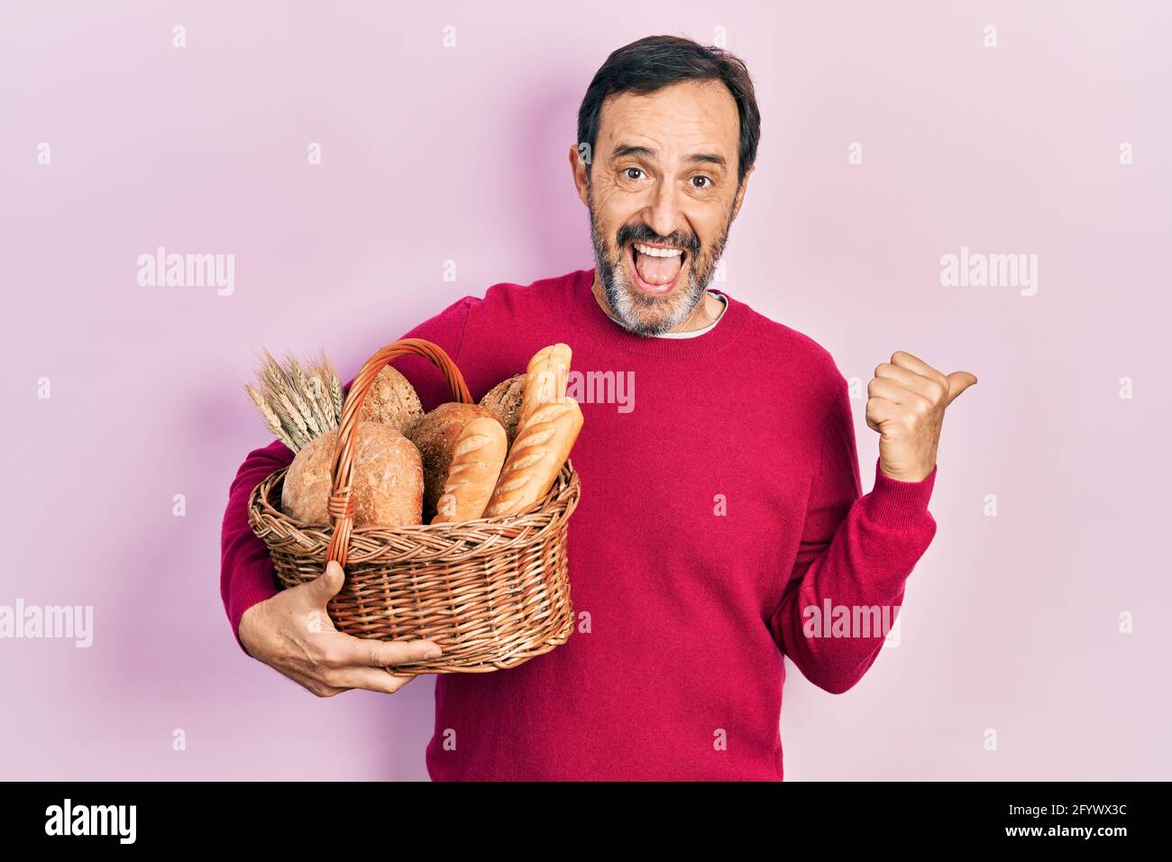 Middle age hispanic man holding wicker basket with bread pointing thumb ...