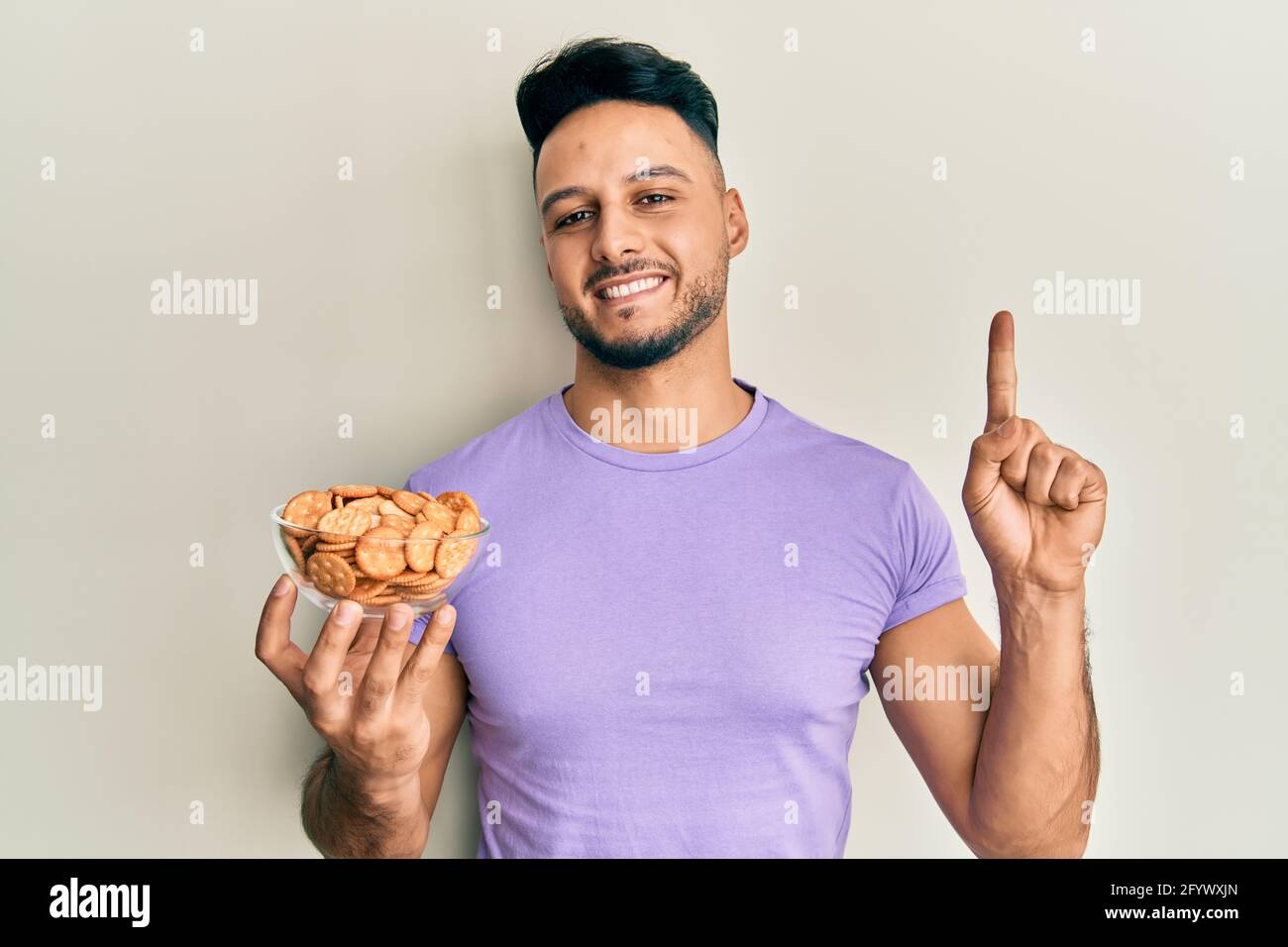Young arab man holding bowl of salty crackers biscuits smiling with an ...