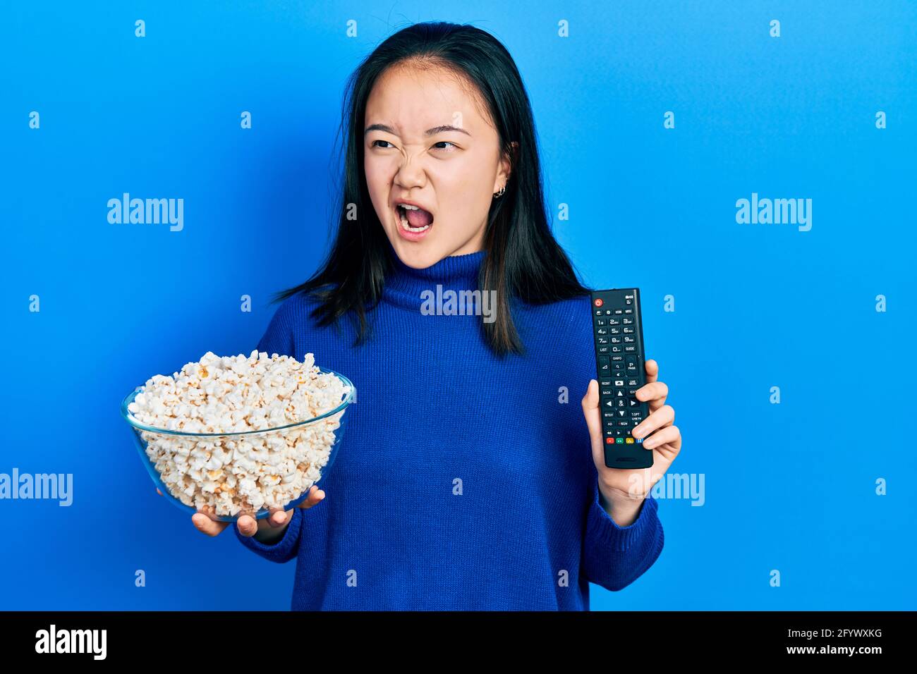 Young chinese girl eating popcorn using tv control angry and mad ...