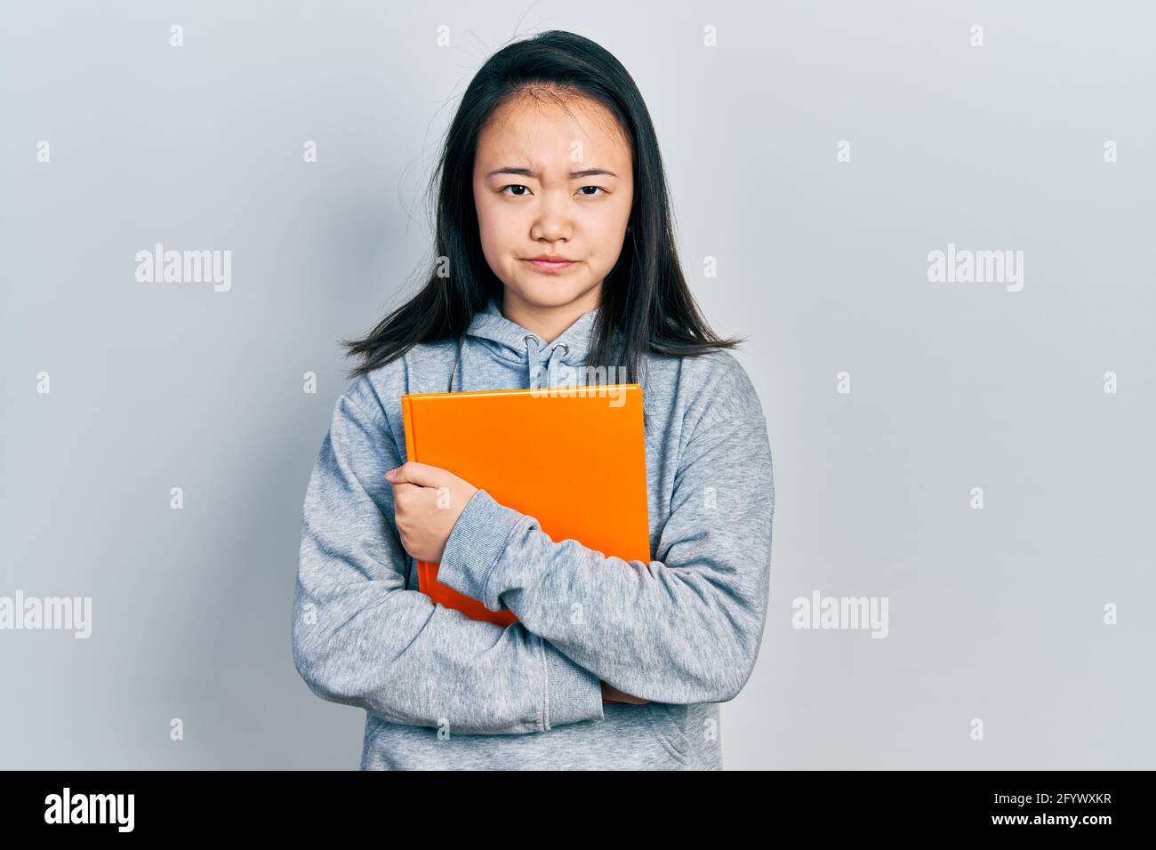 Young chinese girl holding book skeptic and nervous, frowning upset ...