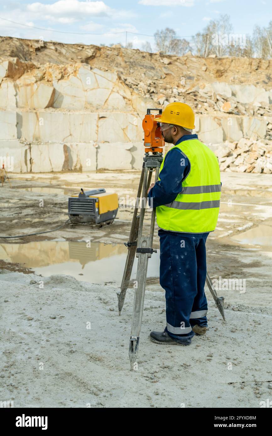 Young surveyor in workwear using geodetic station on construction place ...