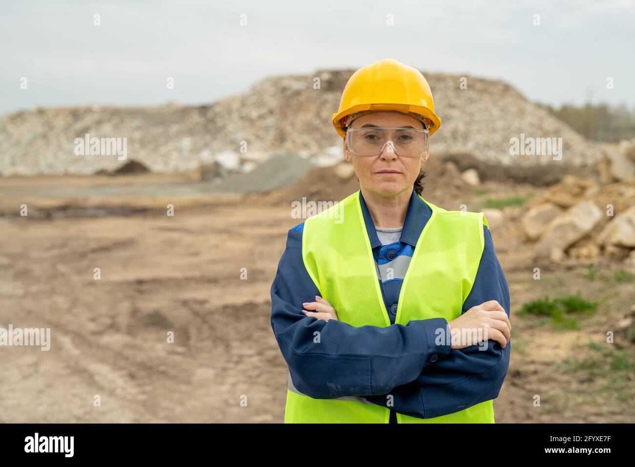 Serious female worker in uniform and protective helmet crossing arms on ...