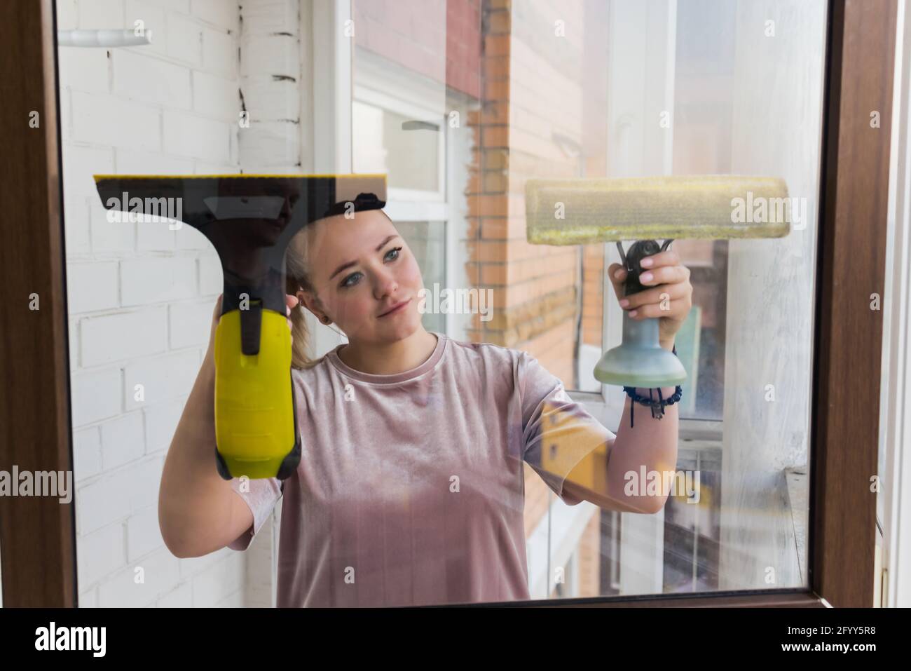 Beautiful young housewife in working clothes washing windows by ...