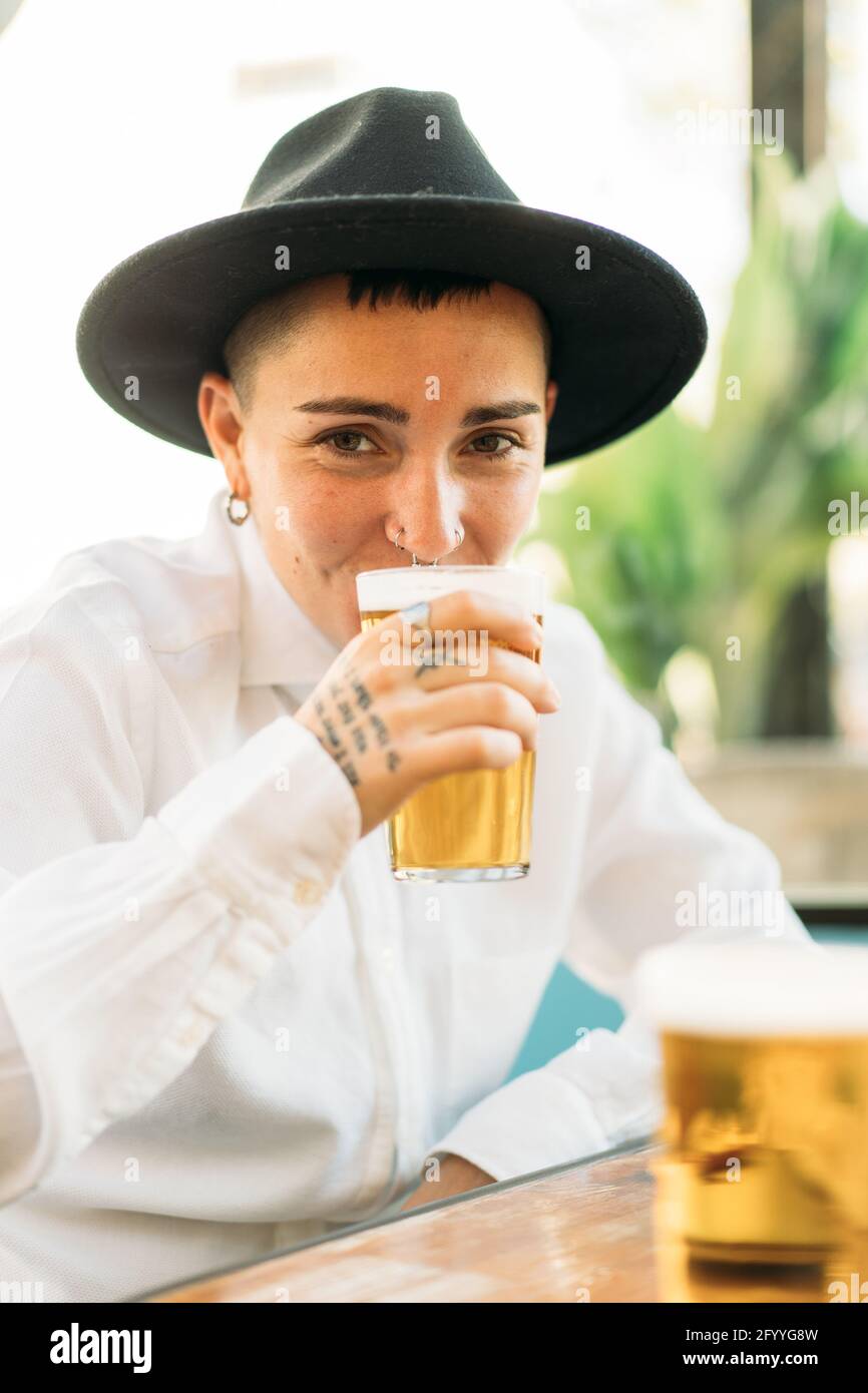 Positive tomboy with tattoos wearing hat and white shirt drinking beer ...