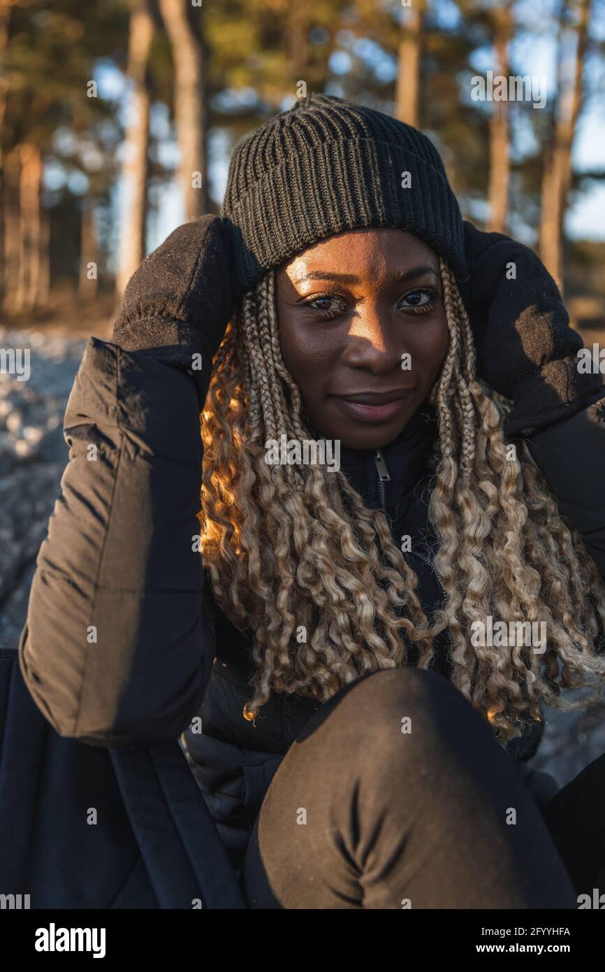Young African American female adjusting hat while sitting against trees ...