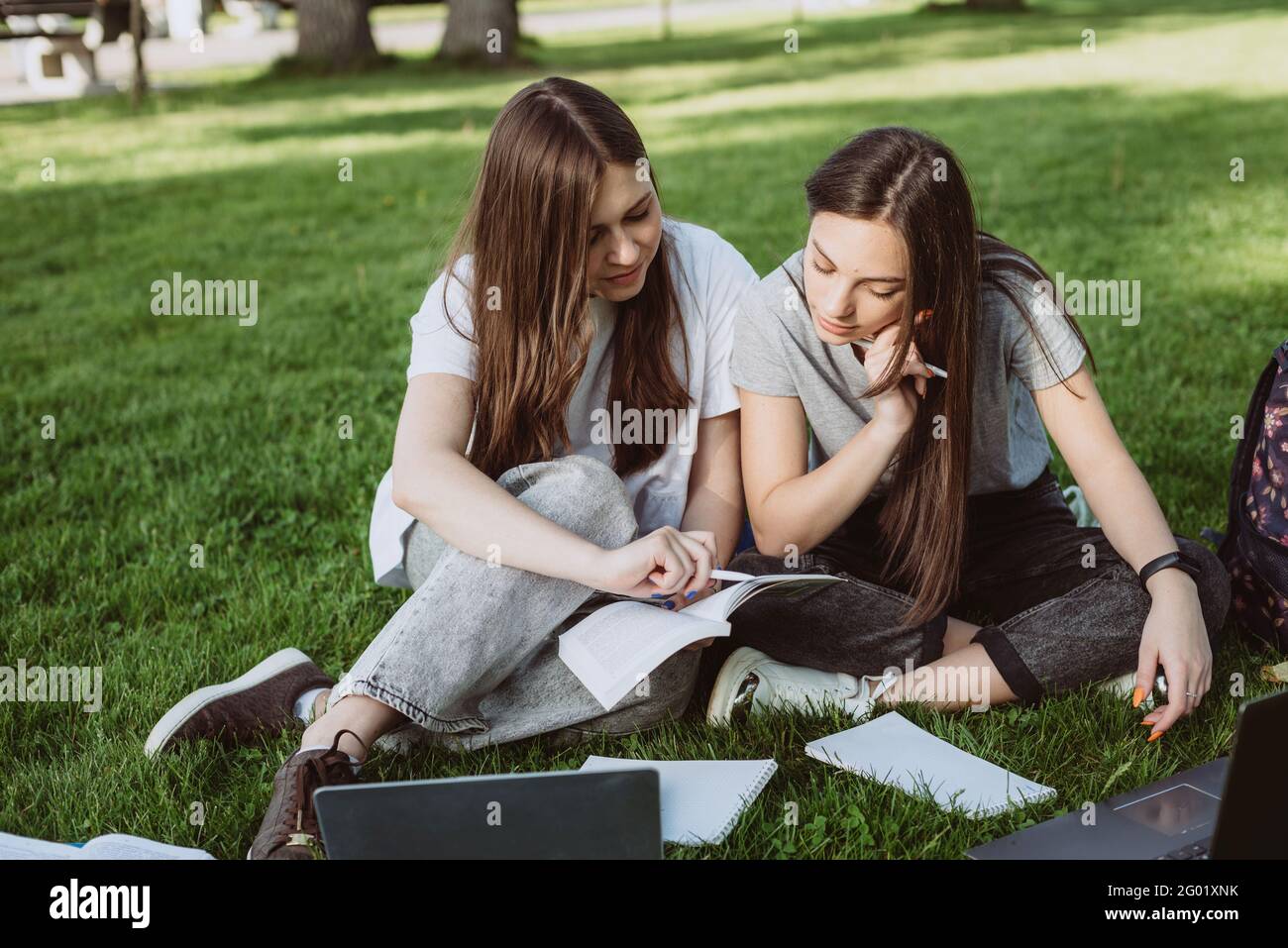 Two female students are sitting in the park on the grass with books and ...