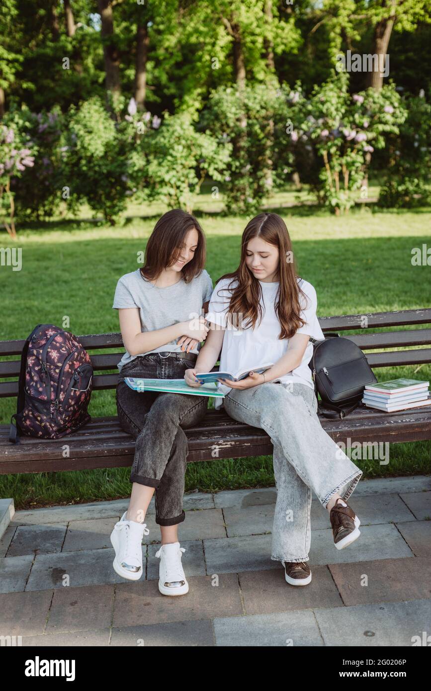 Two student girls are looking at an open book on a bench in the park ...