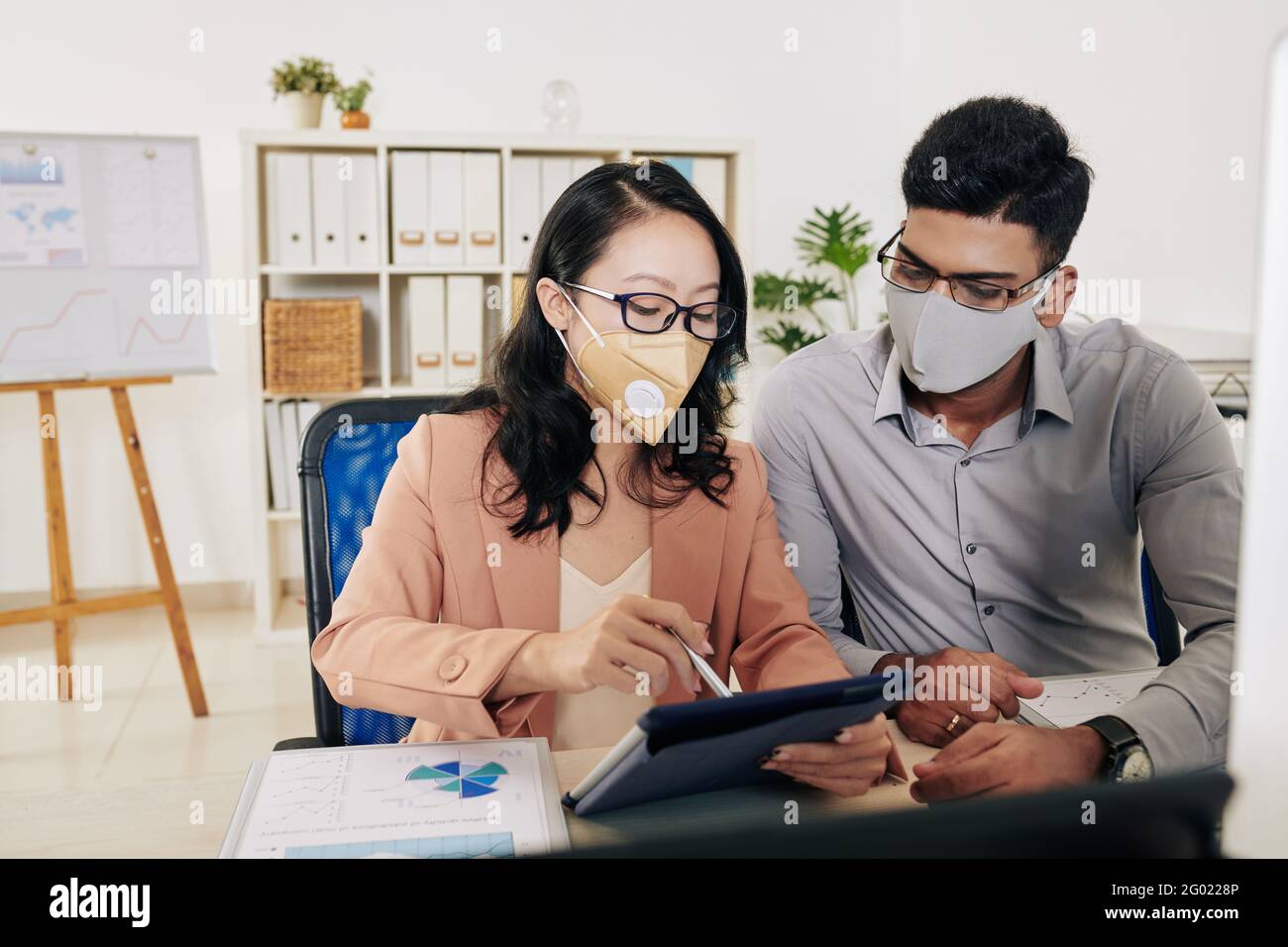 Serious business colleagues in protective masks sitting at office desk ...