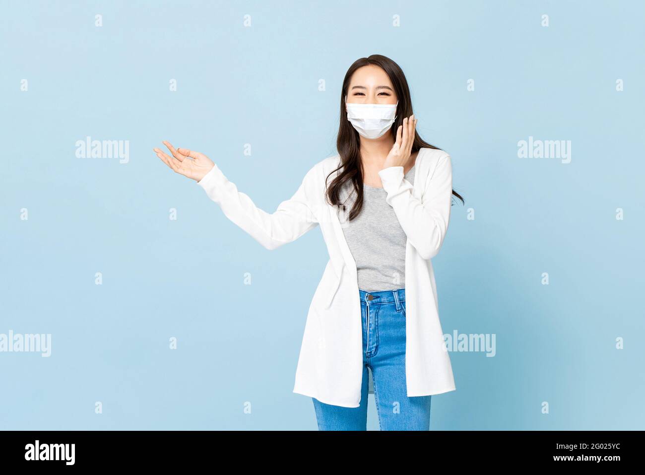 Young Asian woman wearing face mask opening hand to empty space aside ...